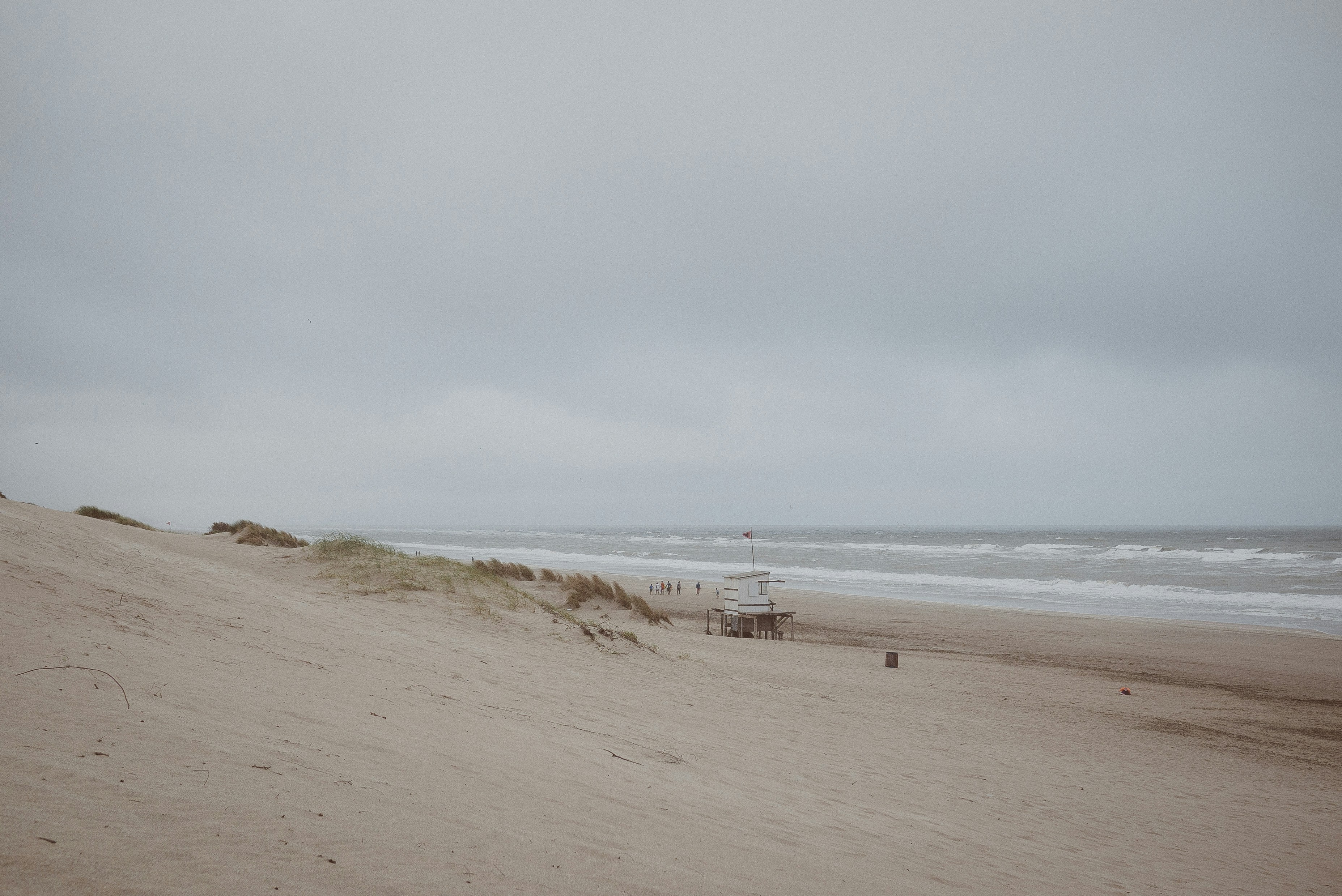 A serene beach scene featuring rolling dunes and a distant lifeguard tower, under an overcast sky. The gentle waves lap at the shore, creating a tranquil atmosphere.