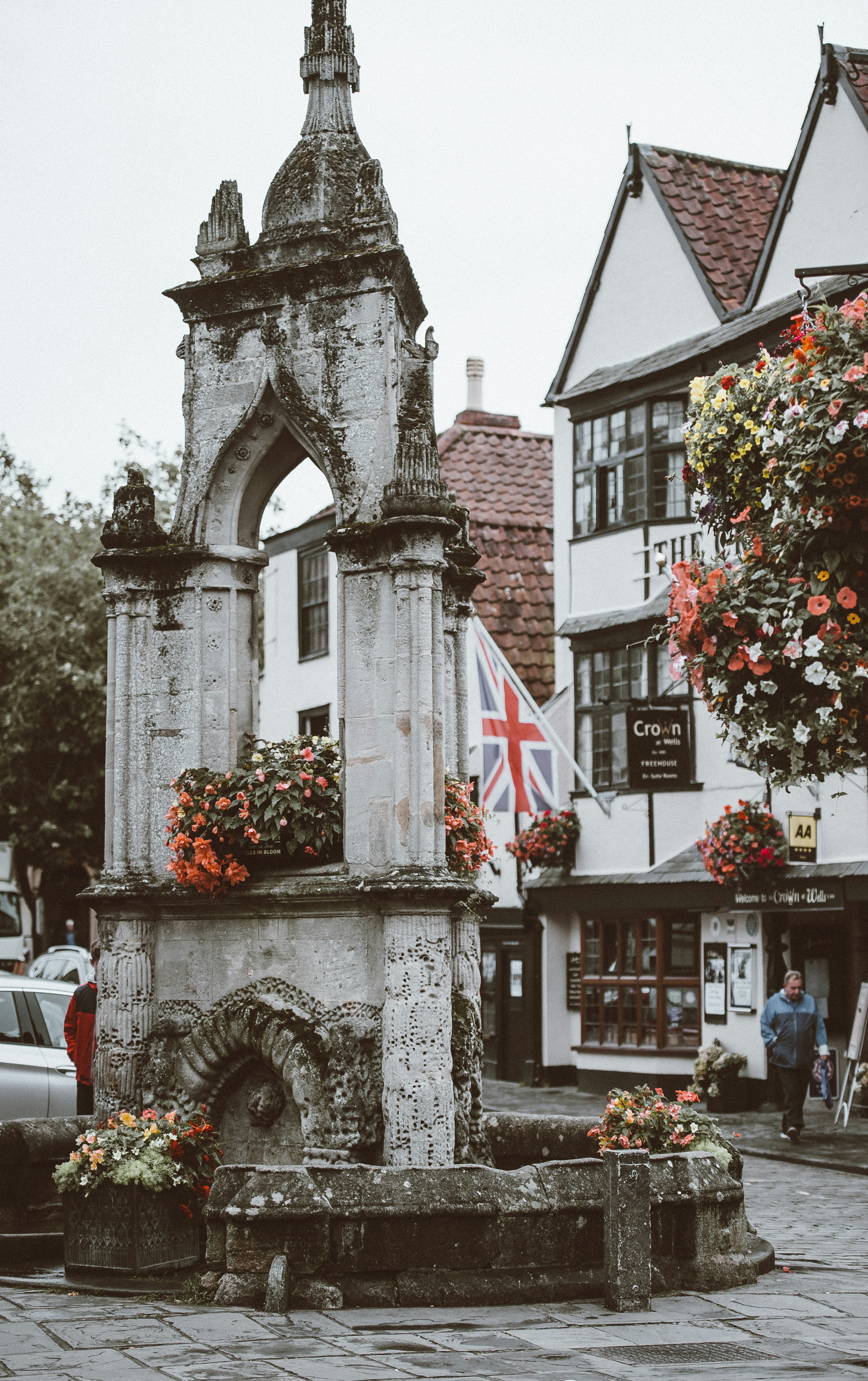 Historic stone clock tower adorned with vibrant flower arrangements, set against traditional English buildings and a Union Jack flag.