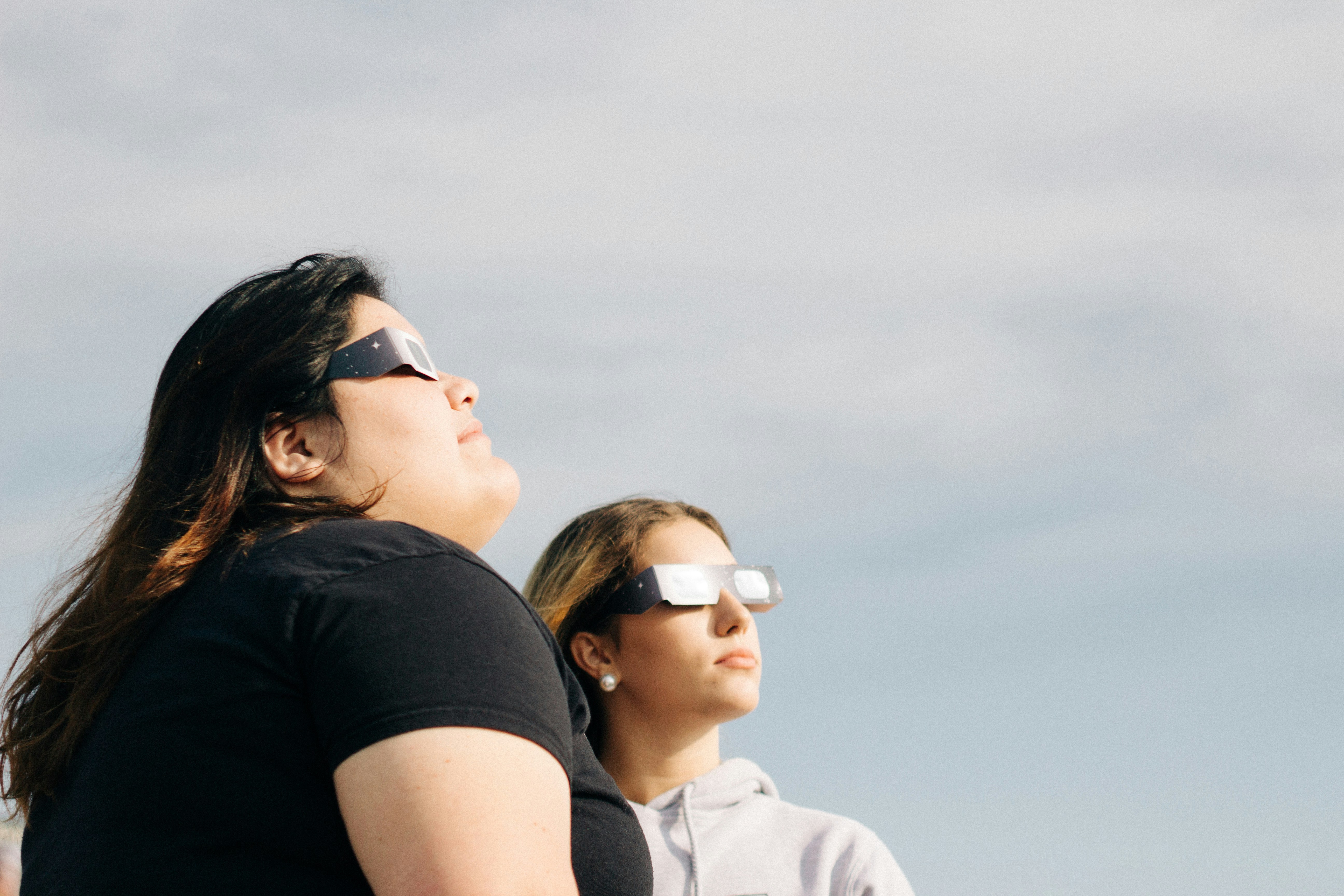 Two individuals wearing protective glasses, looking up at the sky, presumably observing an astronomical event. The scene captures a moment of wonder and anticipation.