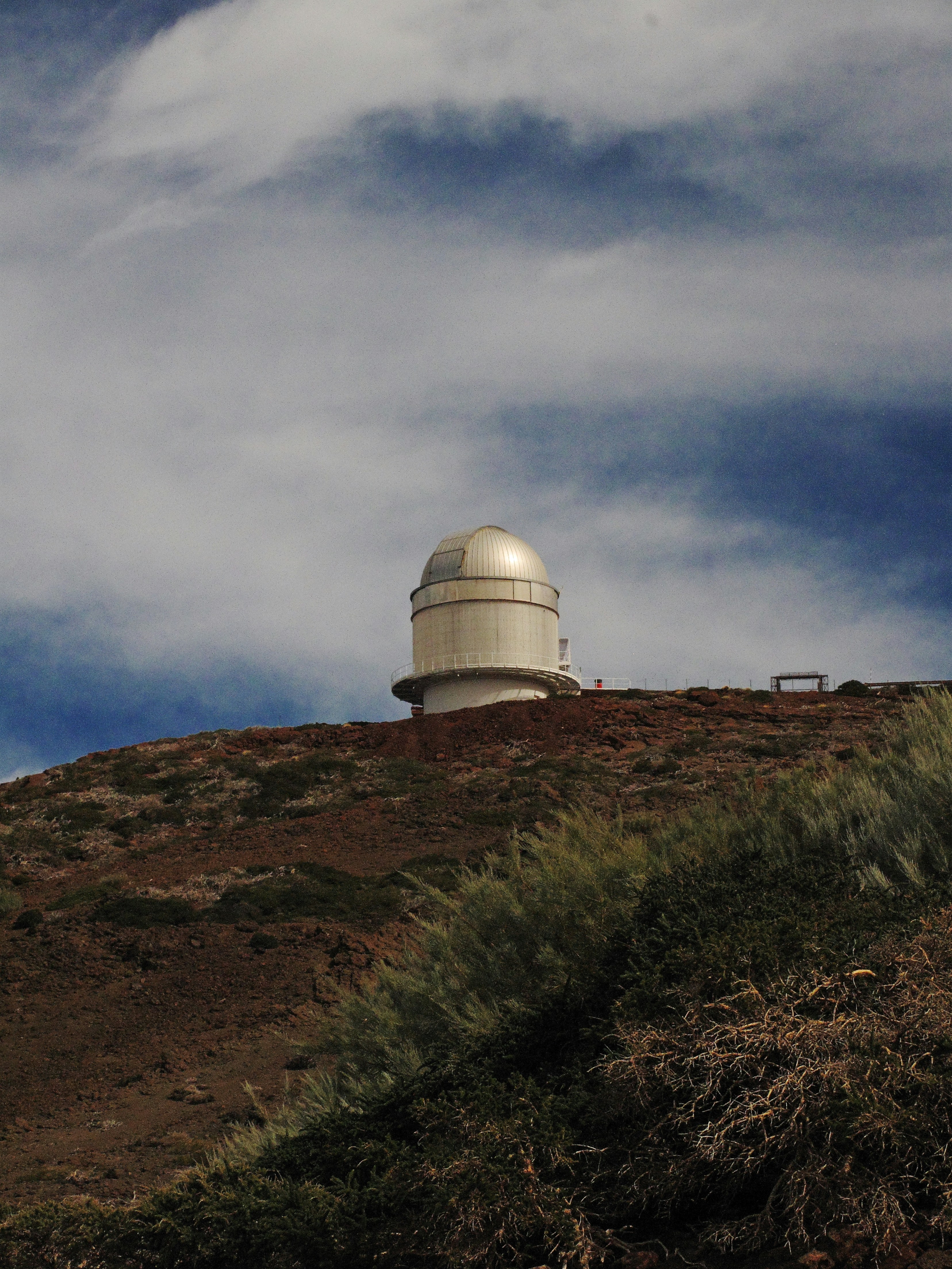 Astronomical observatory perched on a rocky hillside under a vast sky, surrounded by sparse vegetation. The structure stands as a sentinel of the cosmos.