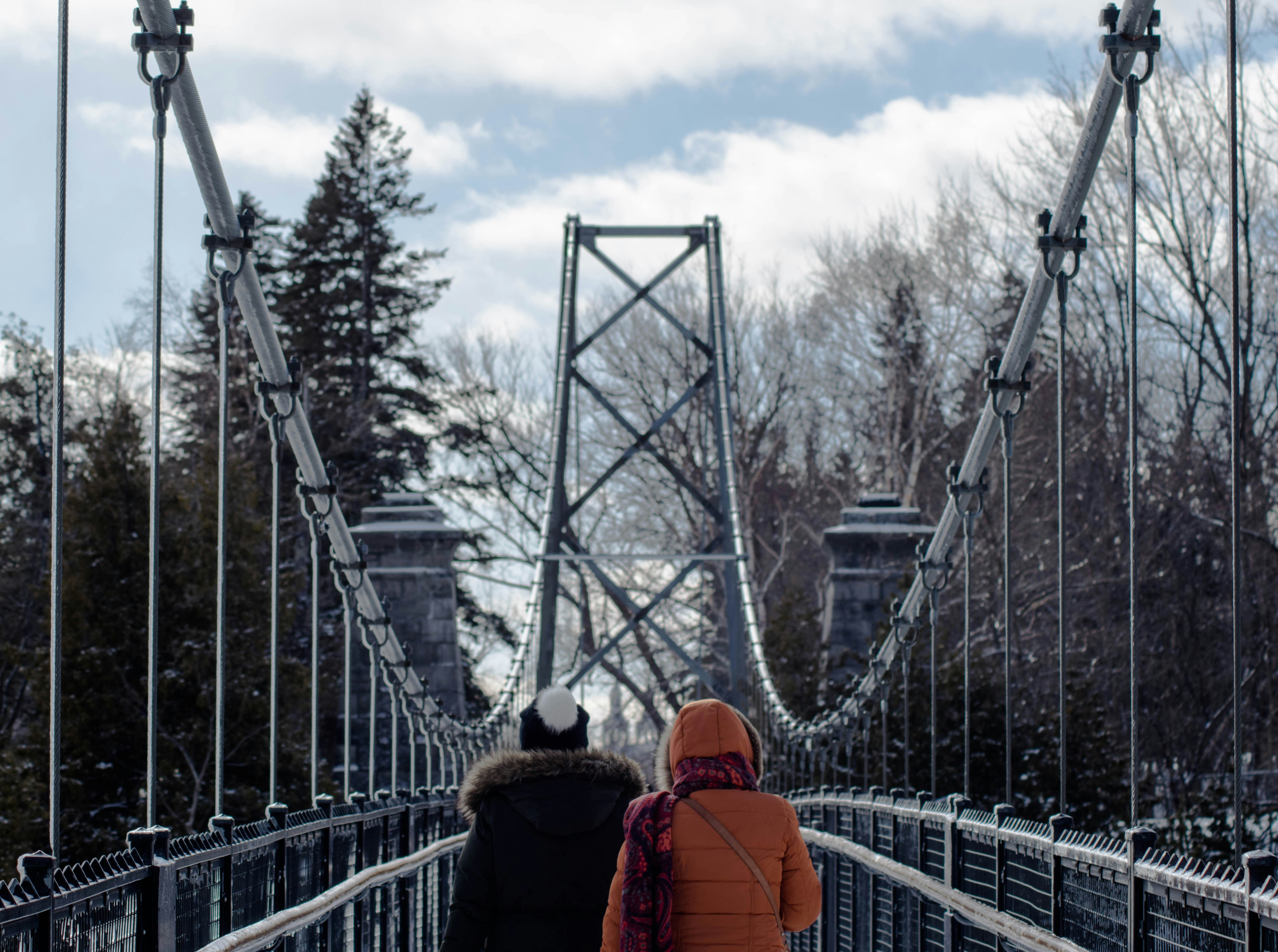 Two people walk on a gray suspension bridge surrounded by winter trees under a cloudy sky.