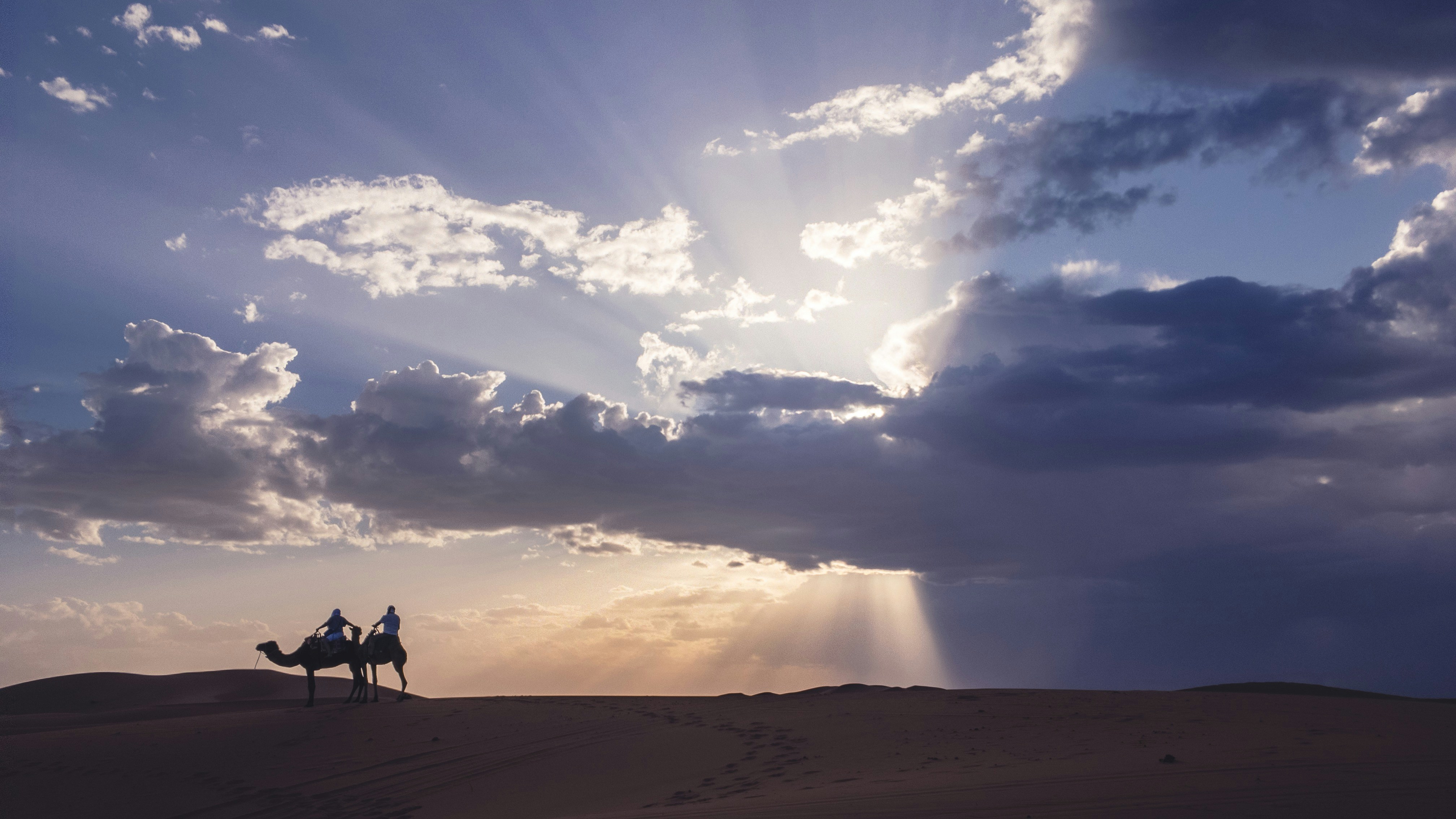 Camel riders silhouetted against a dramatic sky with sun rays piercing through clouds in the Sahara desert.