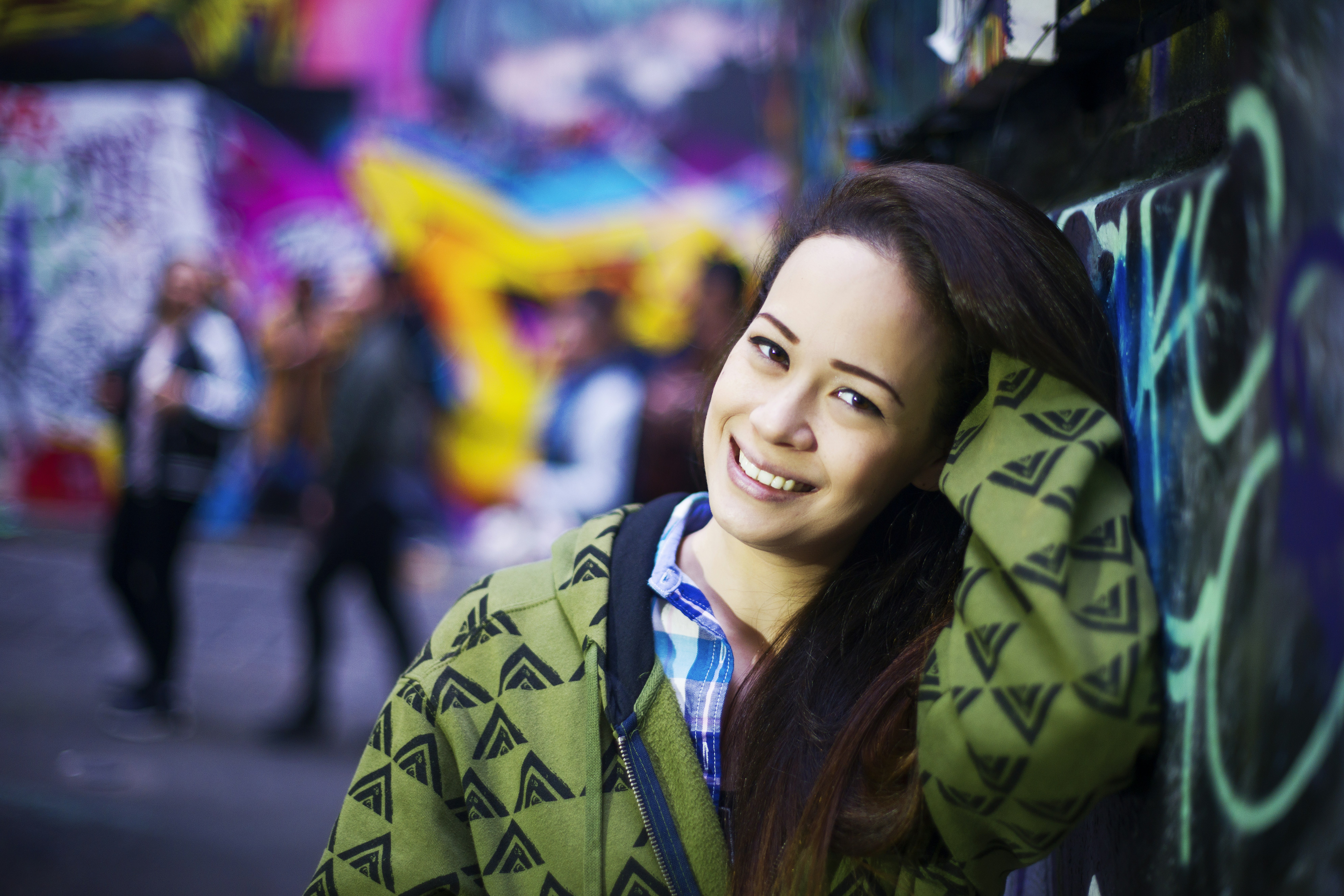 Portrait of a smiling woman leaning against a graffiti-covered wall, with a softly blurred crowd in the background.