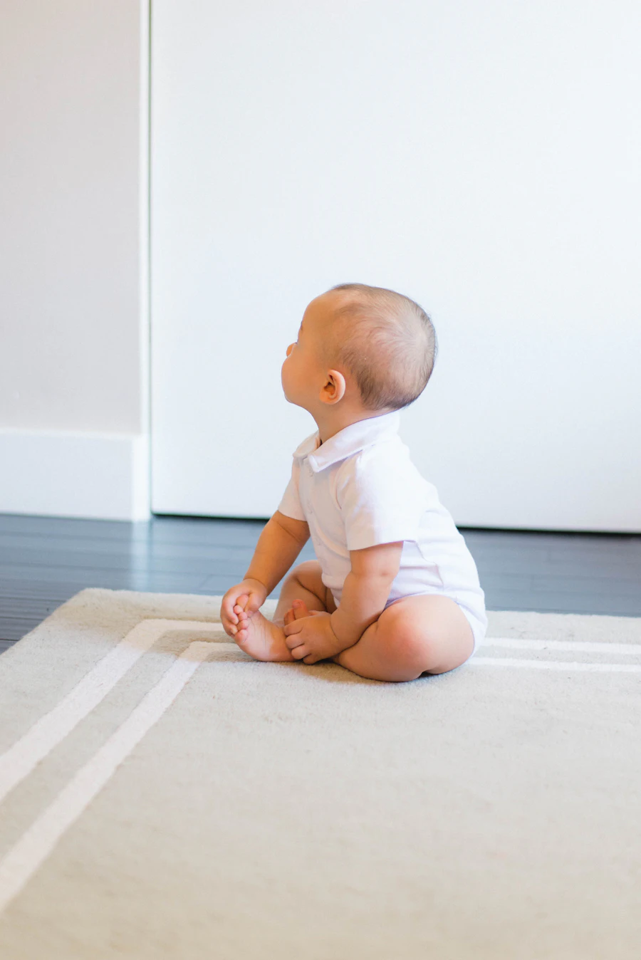 Young child sitting on a soft rug, gazing thoughtfully towards a white door. The simplicity of the scene highlights the innocence of childhood.