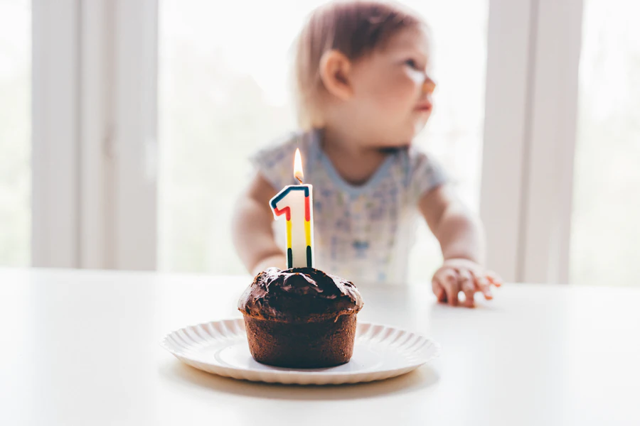 A toddler sits at a bright table with a single-lit cupcake on a plate in the foreground. The candle-lit cupcake is the center of focus while the child remains softly blurred in the background.