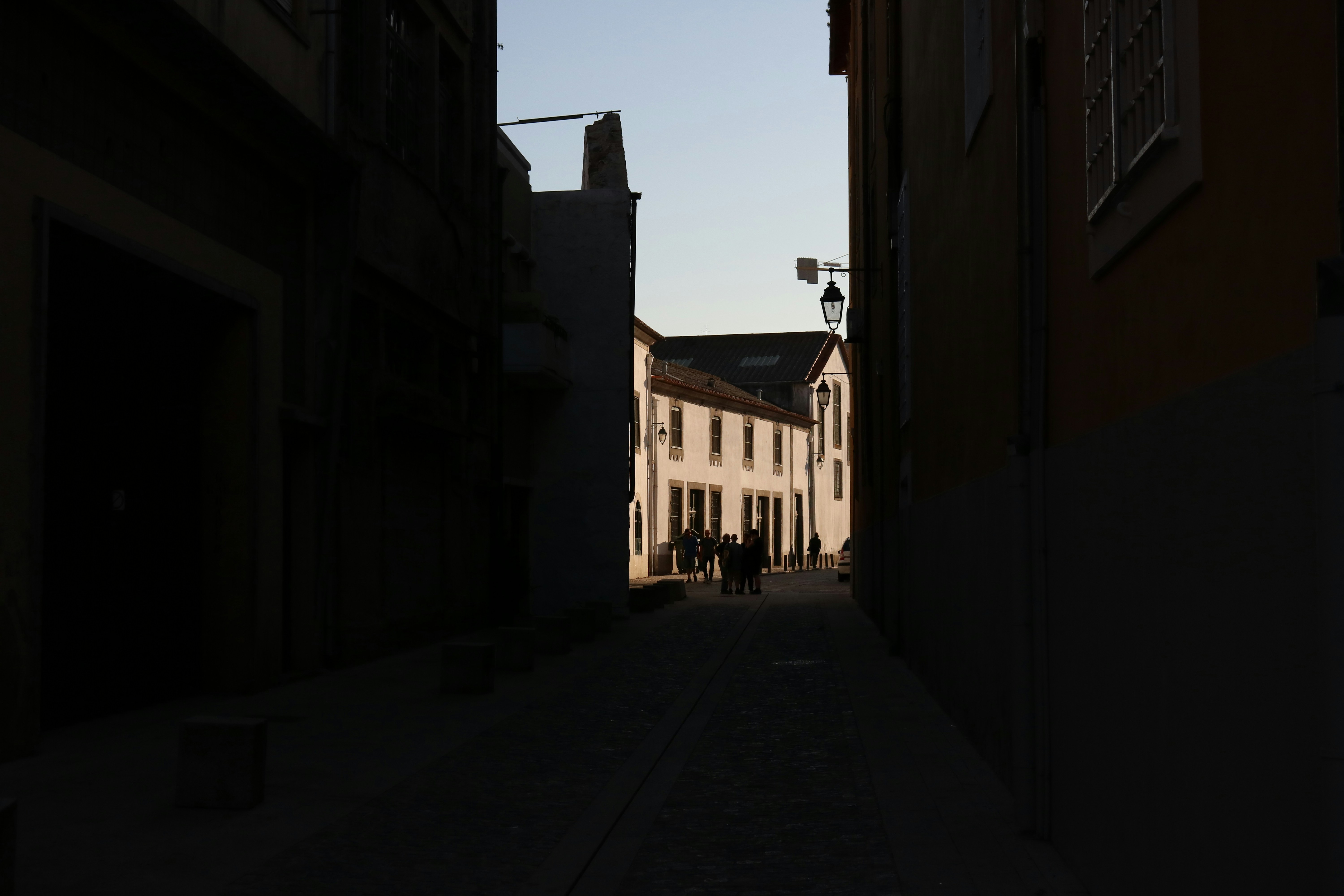 Narrow alleyway leading to sunlit buildings with silhouetted figures walking.