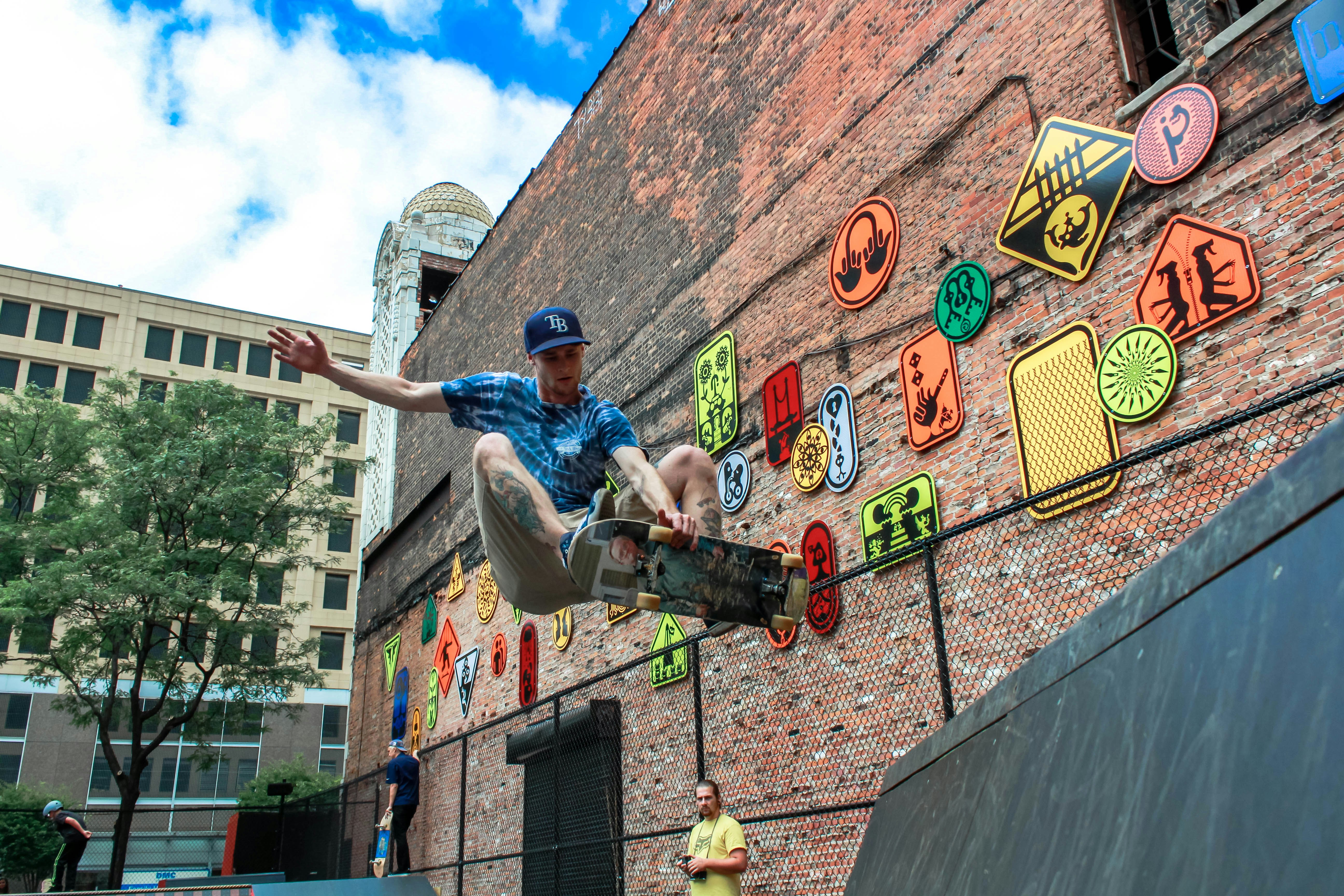Skateboarder performing a trick against a brick wall adorned with colorful art in an urban setting.