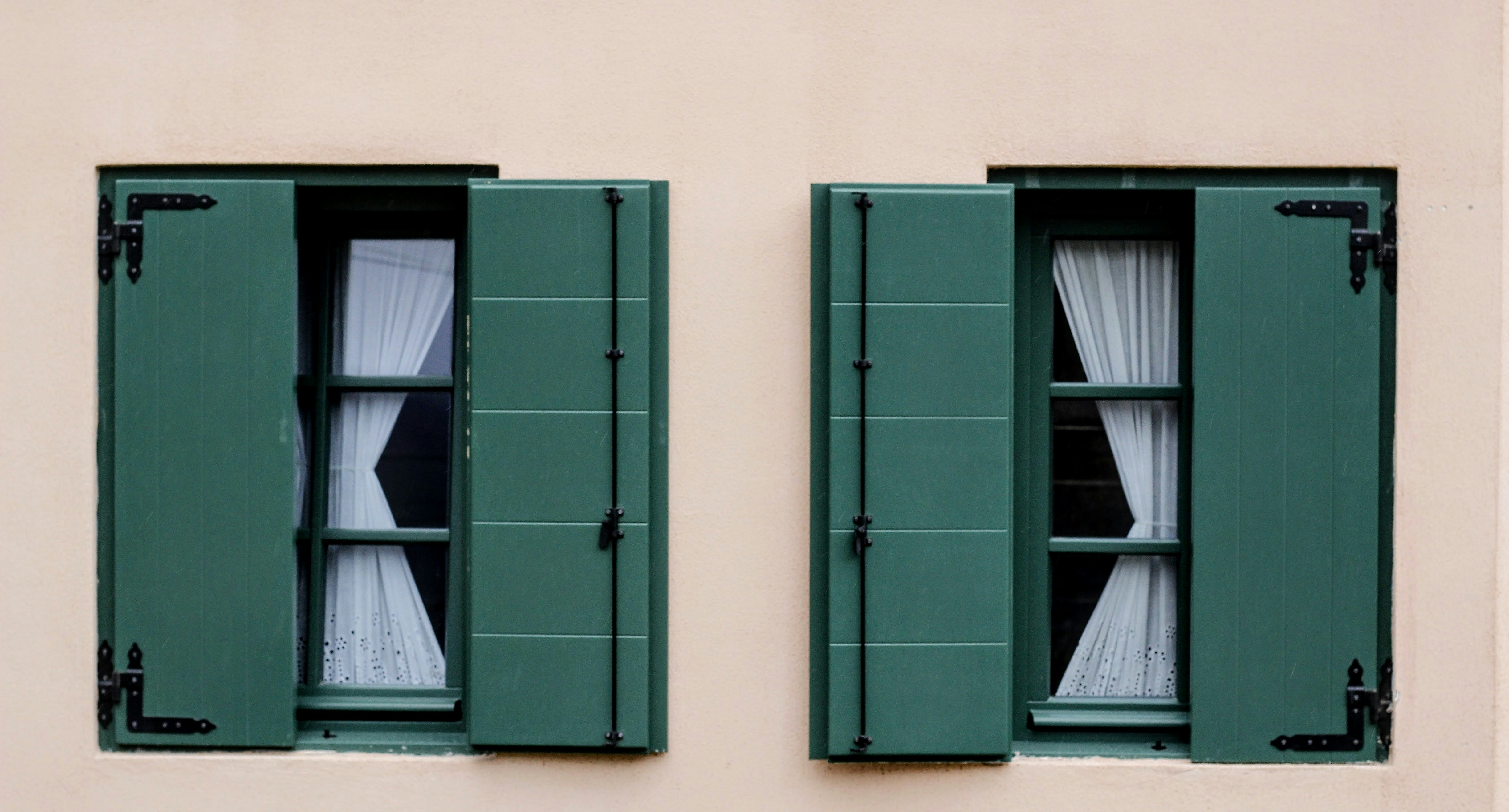 Green wooden shutters on beige wall with white curtains peeking through.