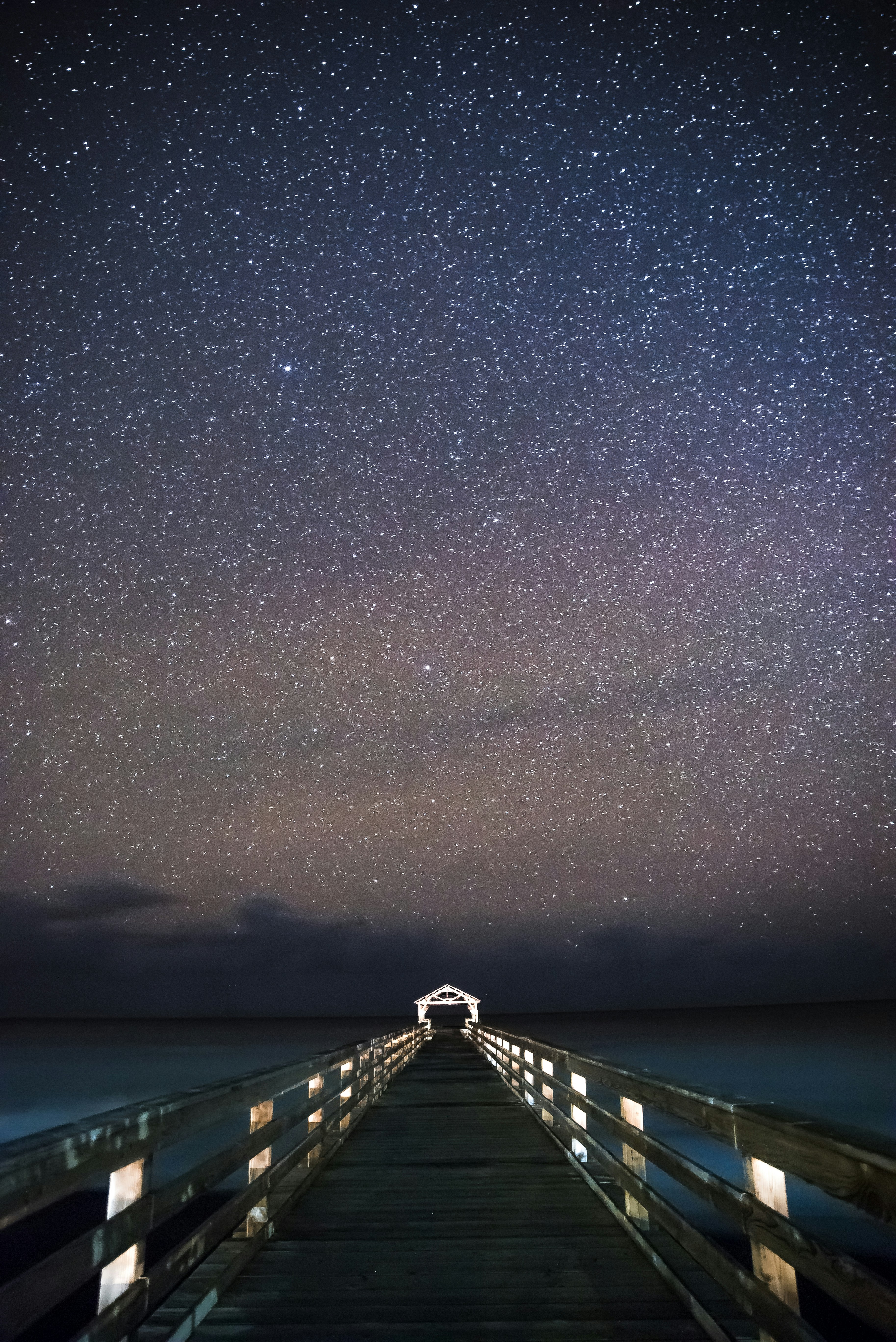 Wooden pier extending into the ocean under a star-filled sky, leading to a brightly lit gazebo at the end.