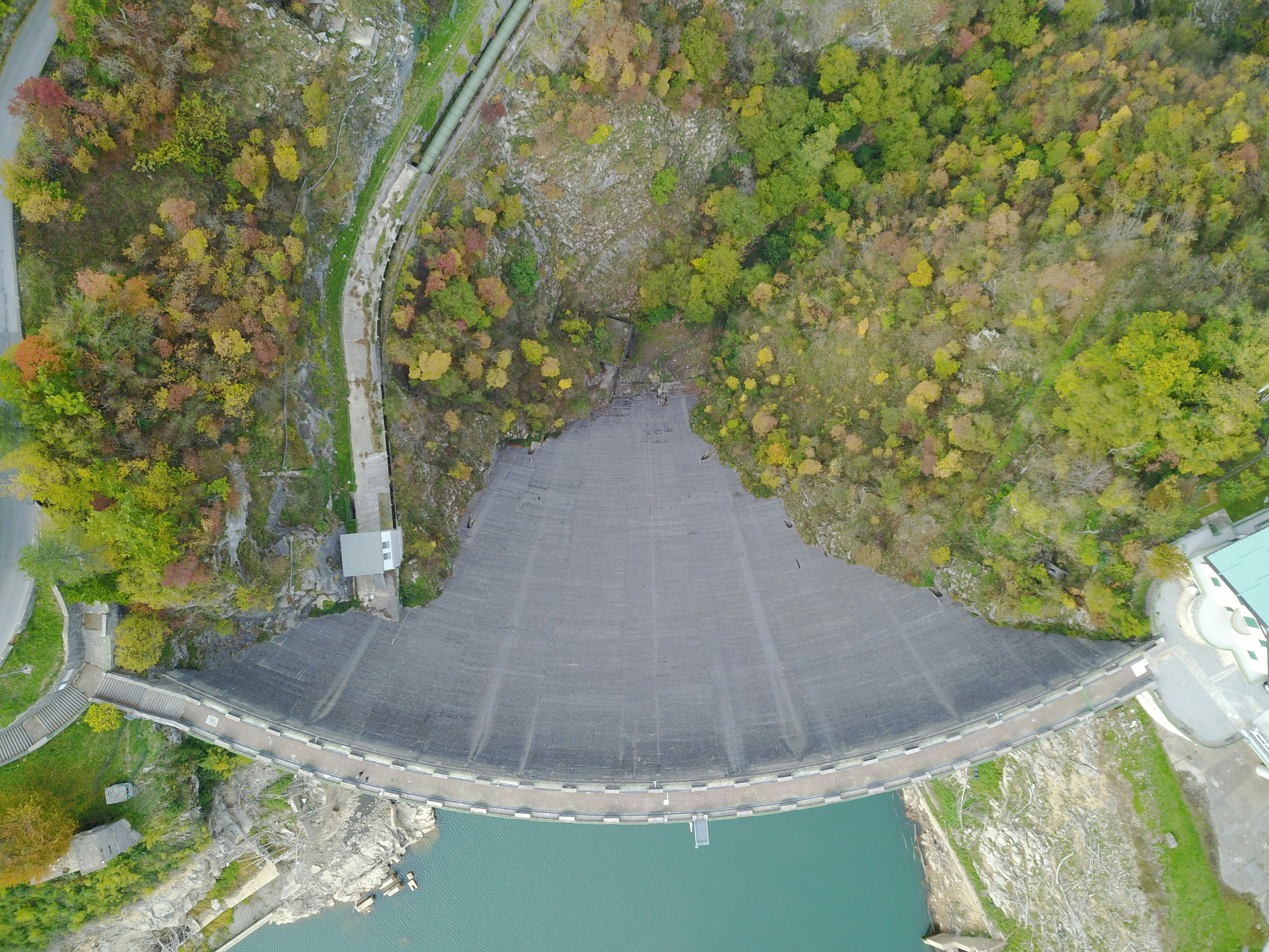 Aerial view showcasing a dam's structure with surrounding foliage in autumn colors. The scene captures the blend of nature and human engineering.