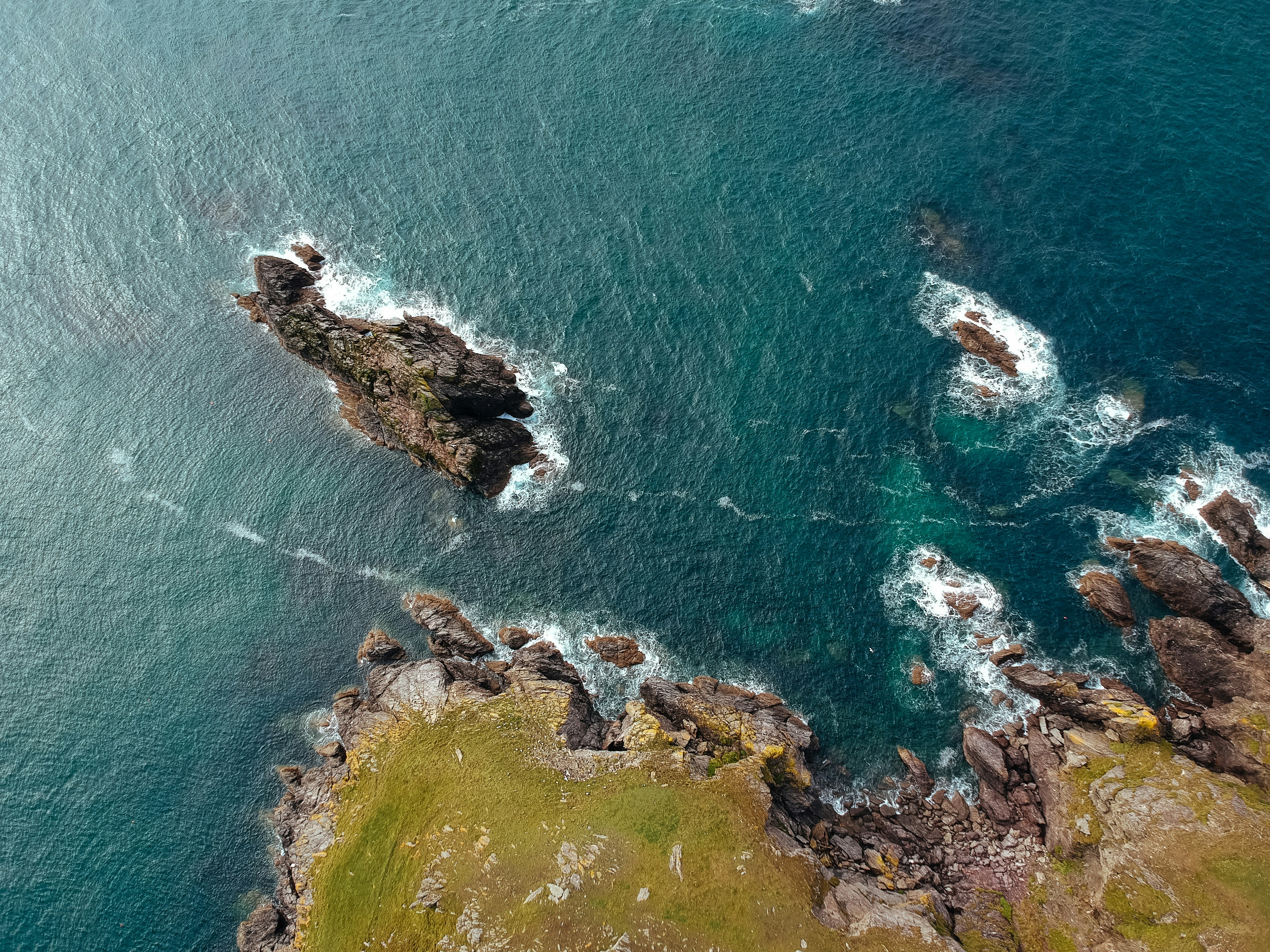 Aerial view of rugged coastline with rocky outcrops surrounded by vibrant blue waters and gentle waves.