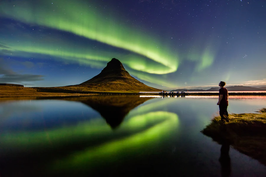 After days of cold, cloud and rain we packed up for our last night in Iceland. We had come to meet Aurora but as many will know it’s not guaranteed. There had been nothing more than a hint of green in the sky for 6 days. But some of the most beautiful things in life happen when we least expect it. As we stared up at this beautiful mountain the sky started to clear and there she was. There’s nothing like it. Watching green waves ripple above you. curving and twisting as if the sky is dancing for you. It’s moments like these that are worth chasing. Worth working for. Worth waiting for. | man standing beside the body of water with Aurora lights in the sky