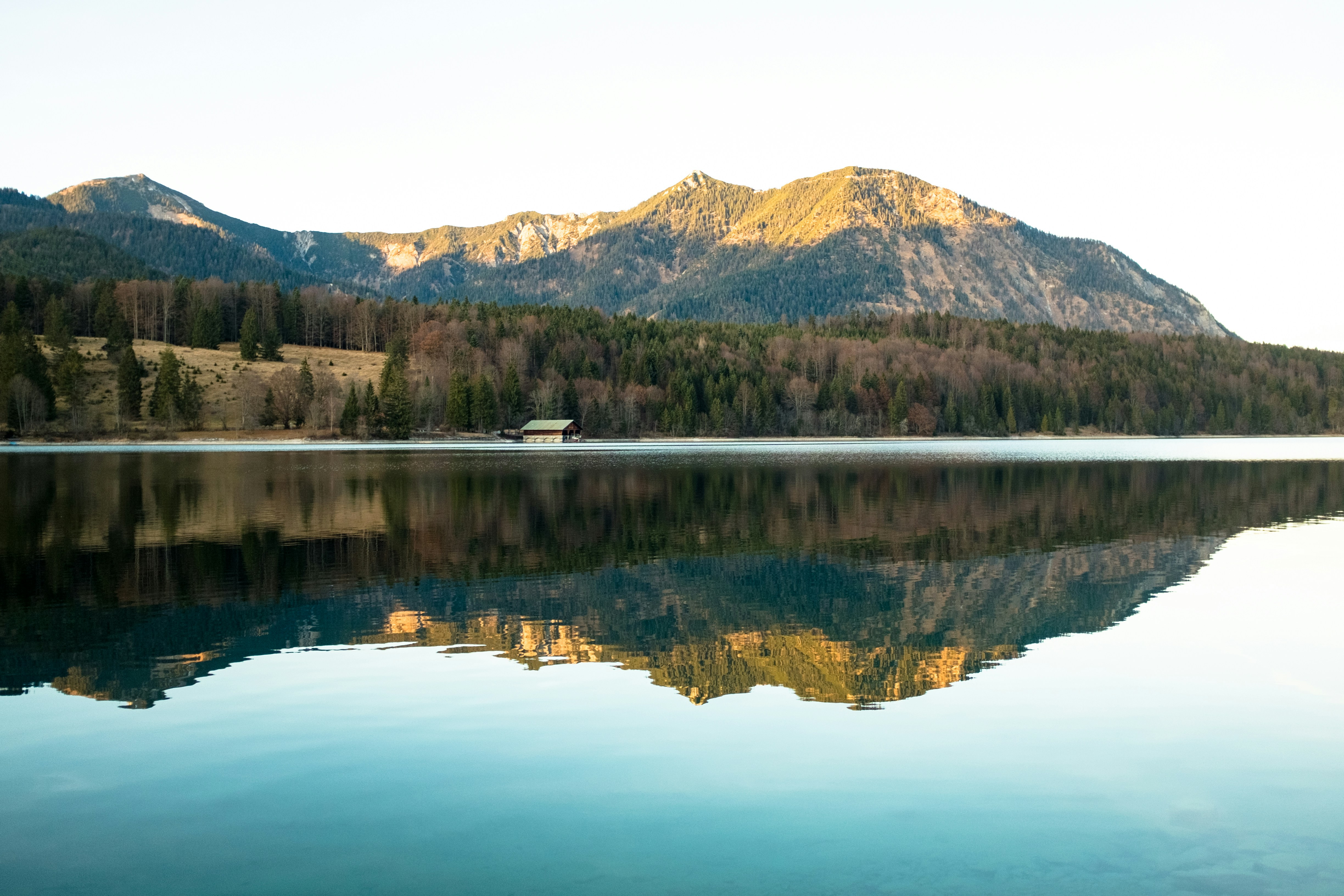 Tranquil lake reflecting majestic mountains under a clear sky, with a rustic cabin nestled among the trees. Perfect harmony between nature and architecture.