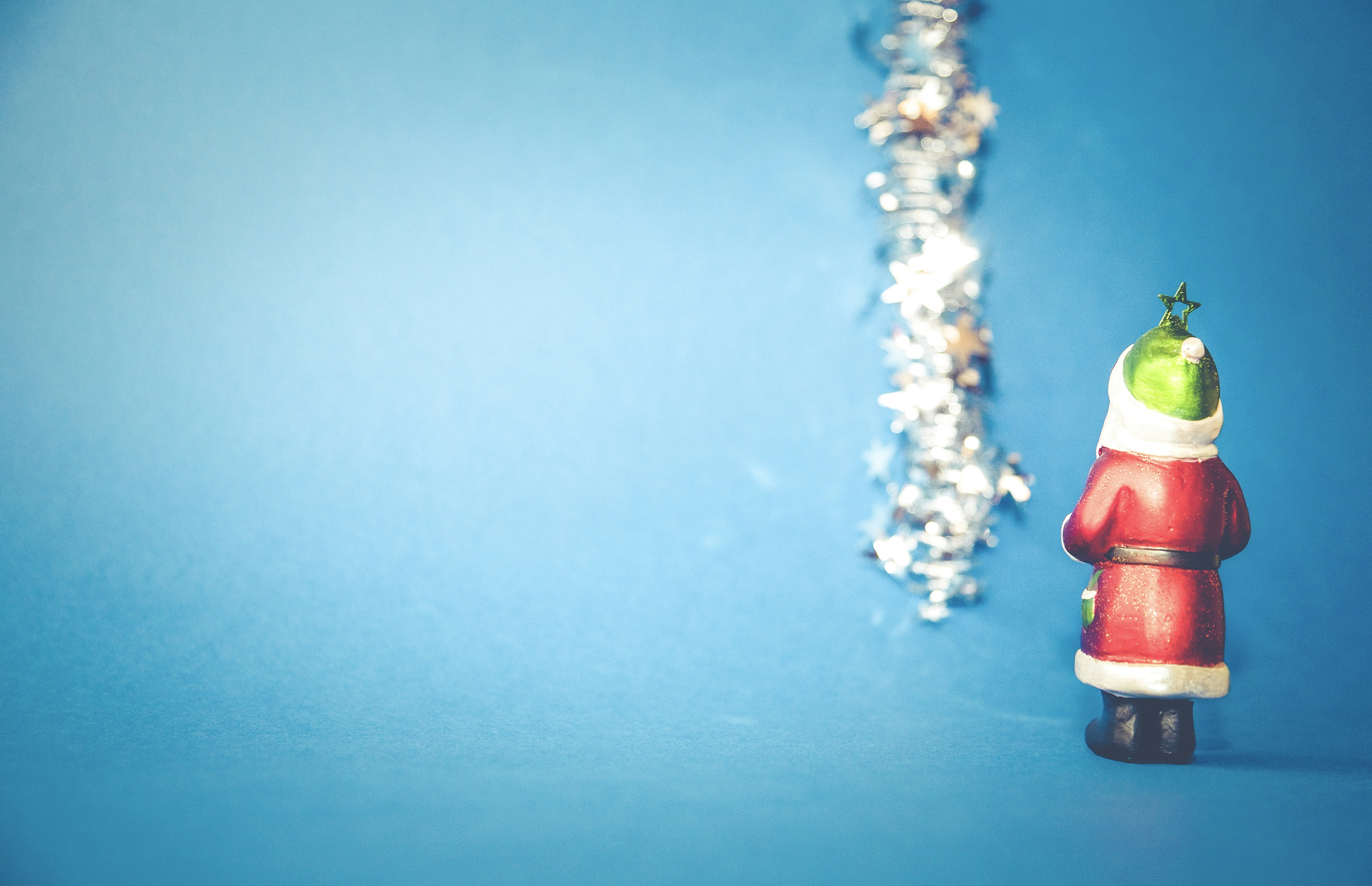 Small Santa Claus figurine in red suit facing silver tinsel on a vibrant blue background.