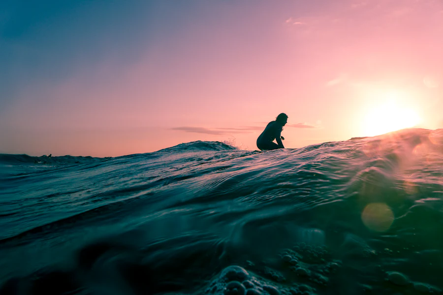 Silhouetted surfer crouches on a wave beneath a glowing pink-orange sunset, with rippling water in the foreground.