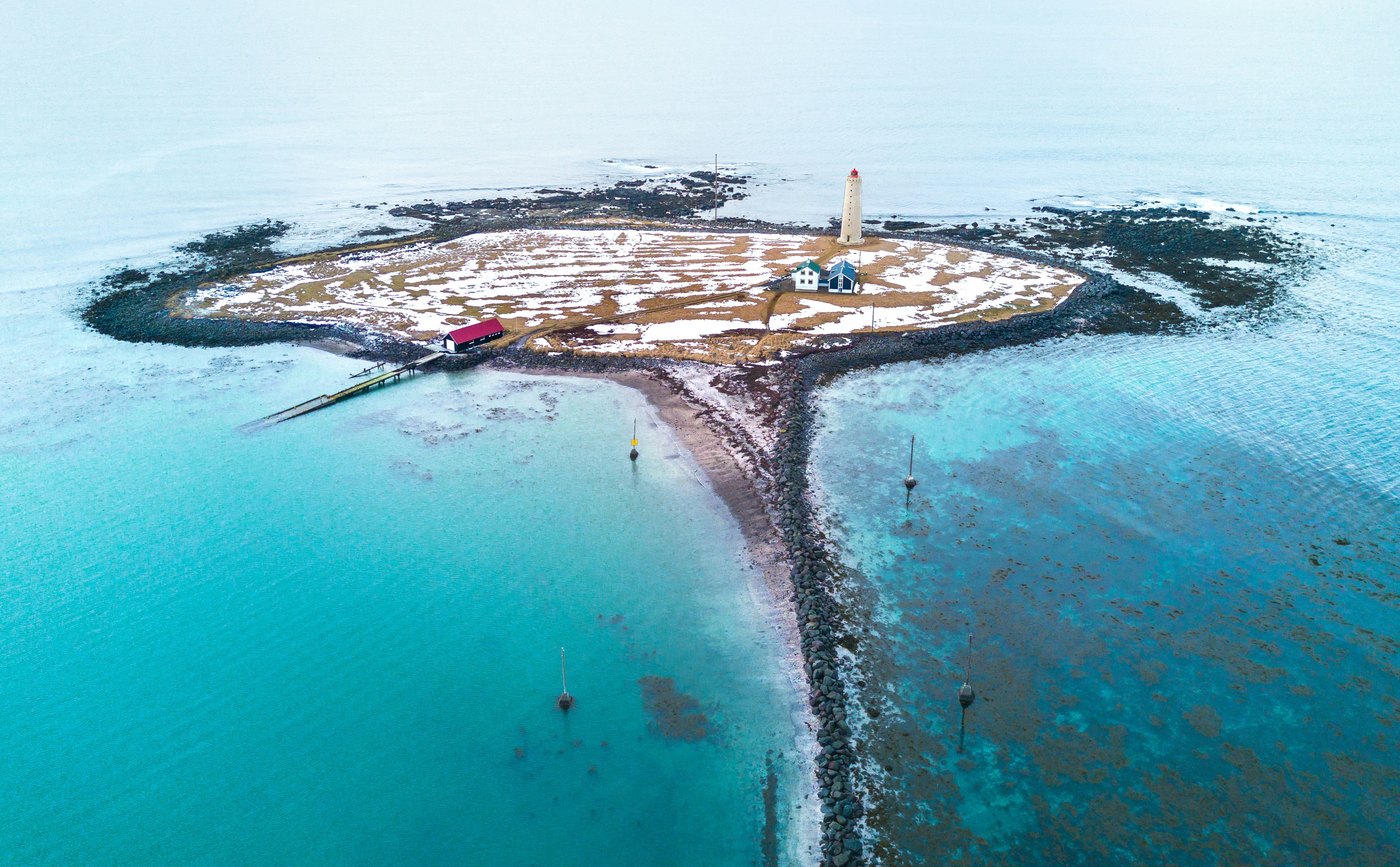 Lighthouse standing on a rocky island surrounded by tranquil turquoise waters, with a quaint house and a red structure nearby. Snow patches dot the landscape.
