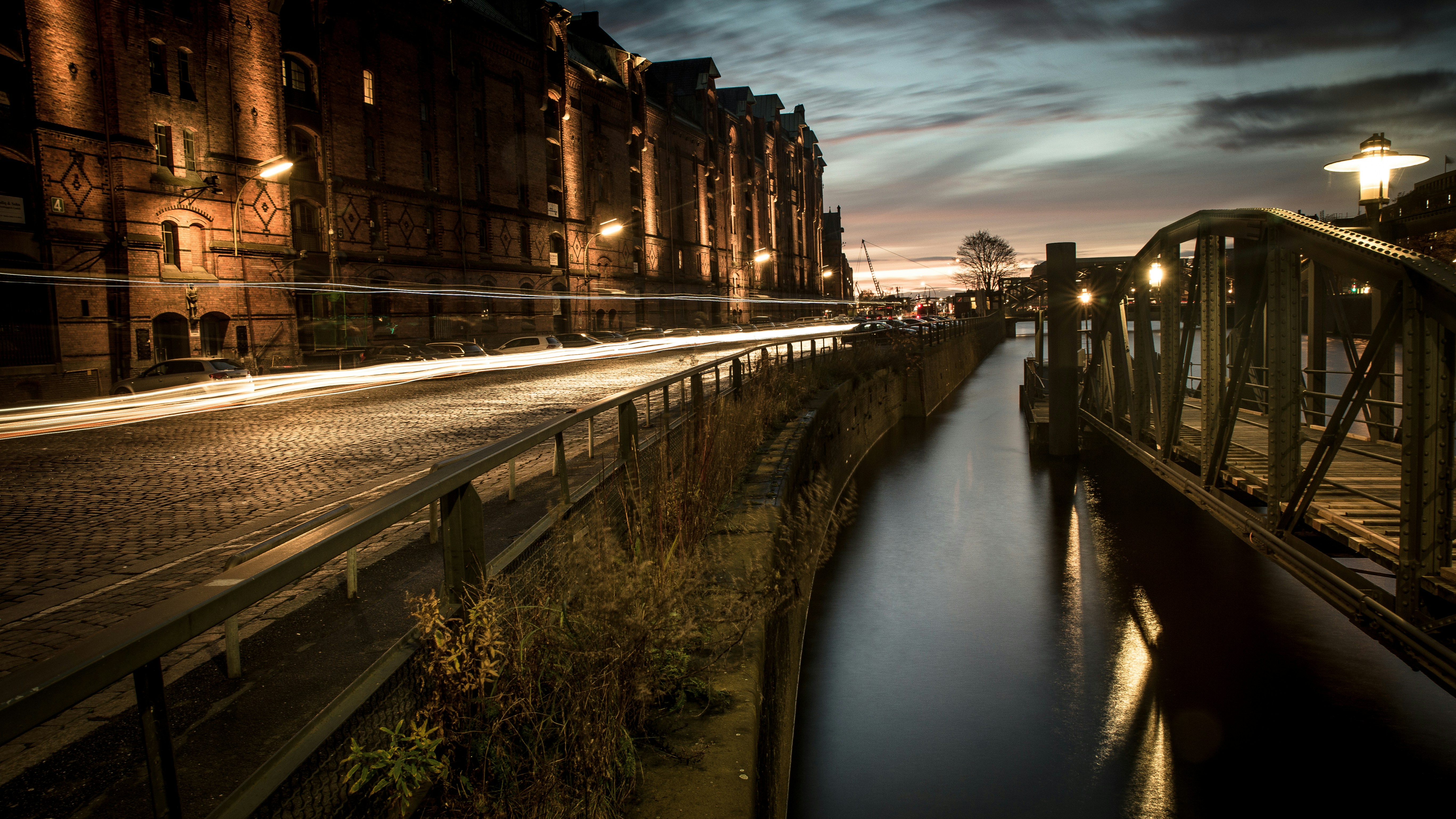 Historic warehouse illuminated by streetlights alongside a tranquil canal, with light trails from passing vehicles. 