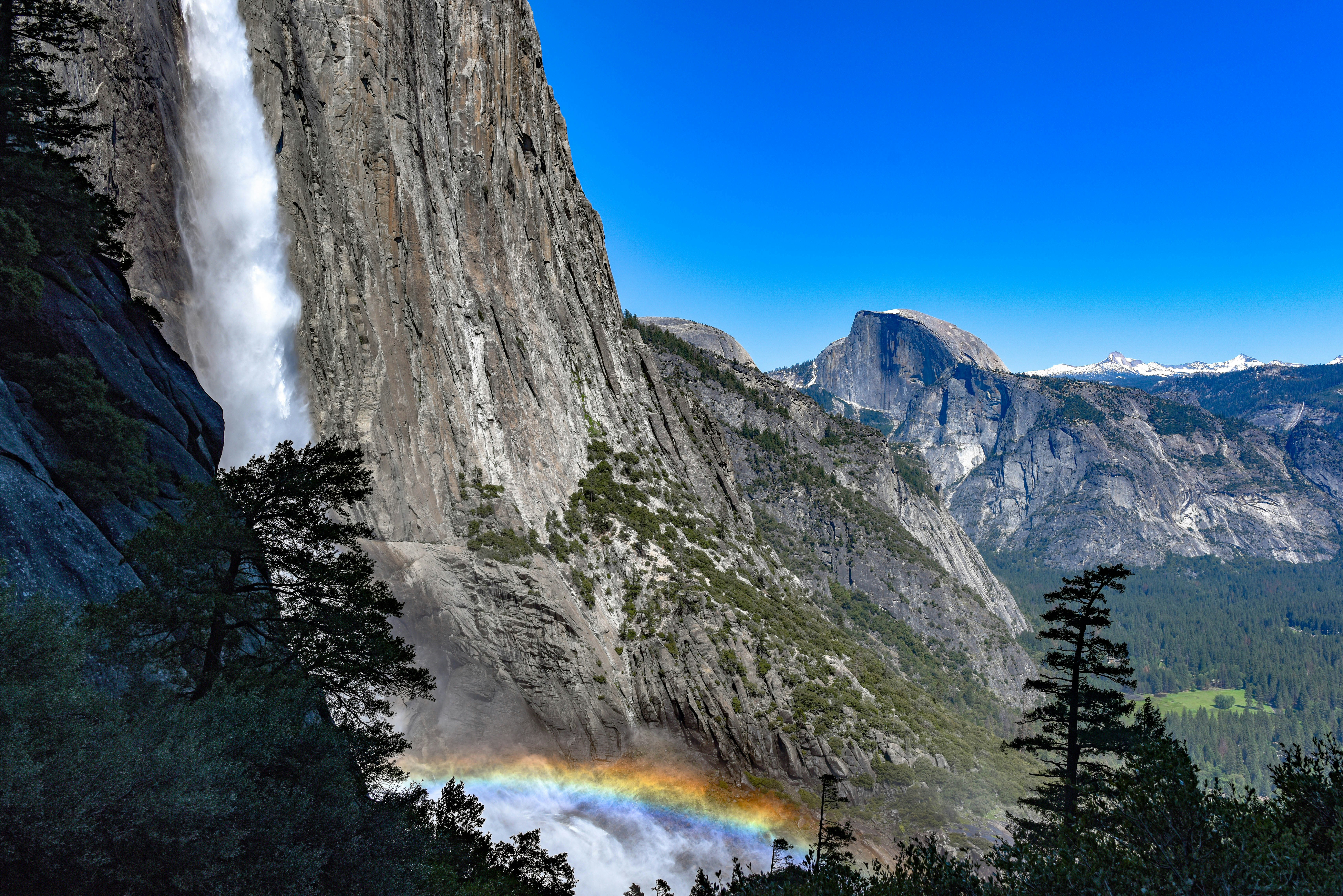Yosemite Falls cascading over granite cliffs with a vibrant rainbow at the base and Half Dome in the background.