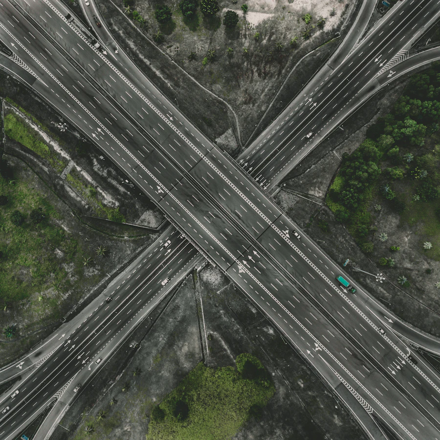 Top-down aerial view of a multi-lane highway interchange crossing in an X shape, surrounded by sparse greenery and gray earth. A green truck is visible on the right side of the overpass.