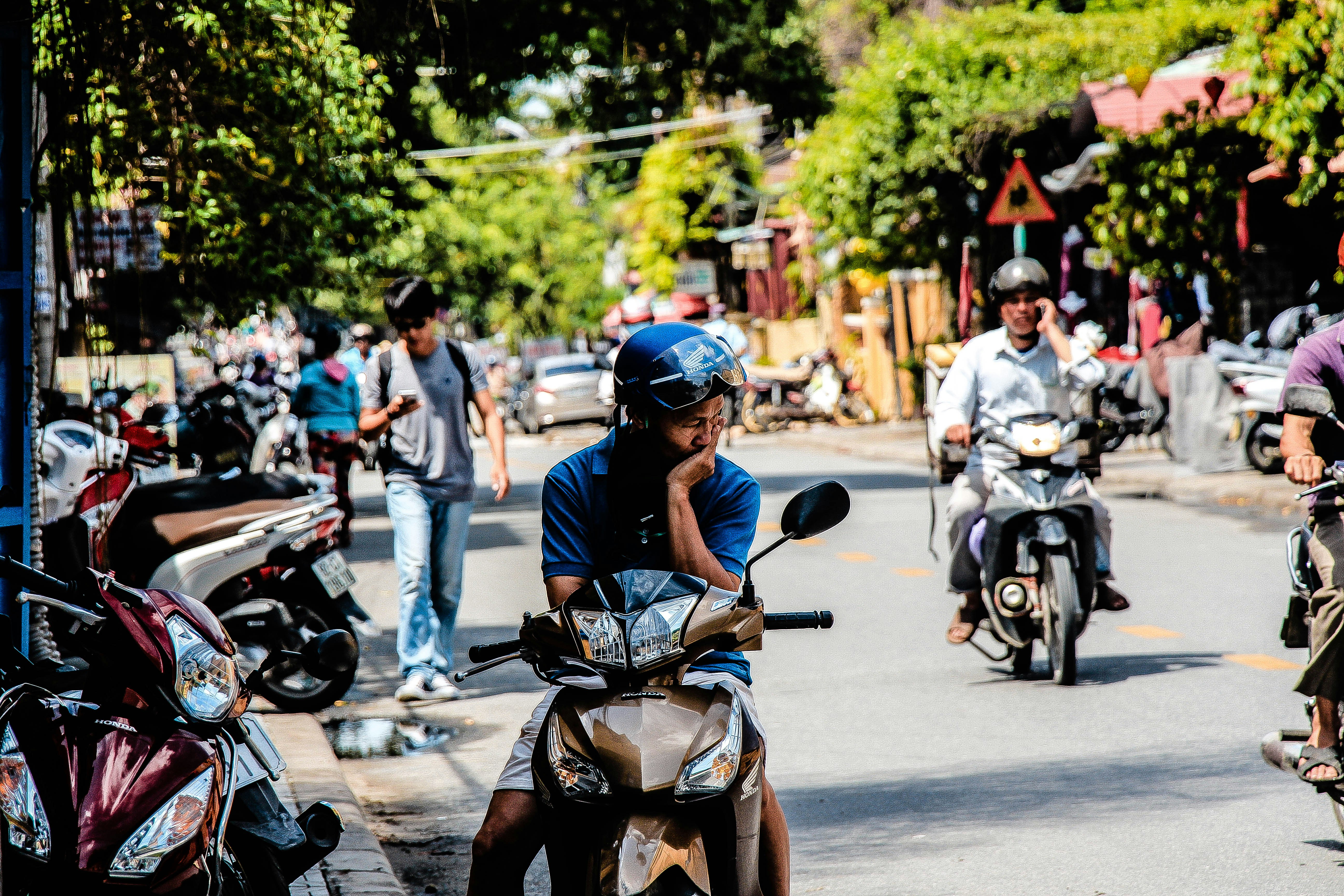 A young man sits pensively on his scooter amidst a bustling street scene filled with motorcycles and pedestrians. The vibrant atmosphere captures the essence of urban life.