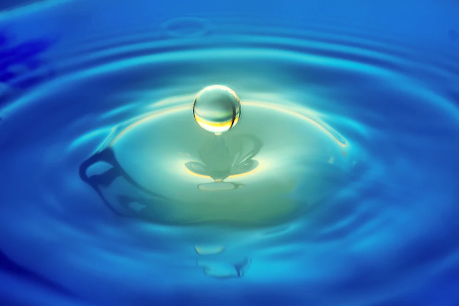 A single water droplet hovers above a glowing pool, sending smooth concentric ripples across a blue surface. The droplet reflects the light in a glassy, abstract scene.