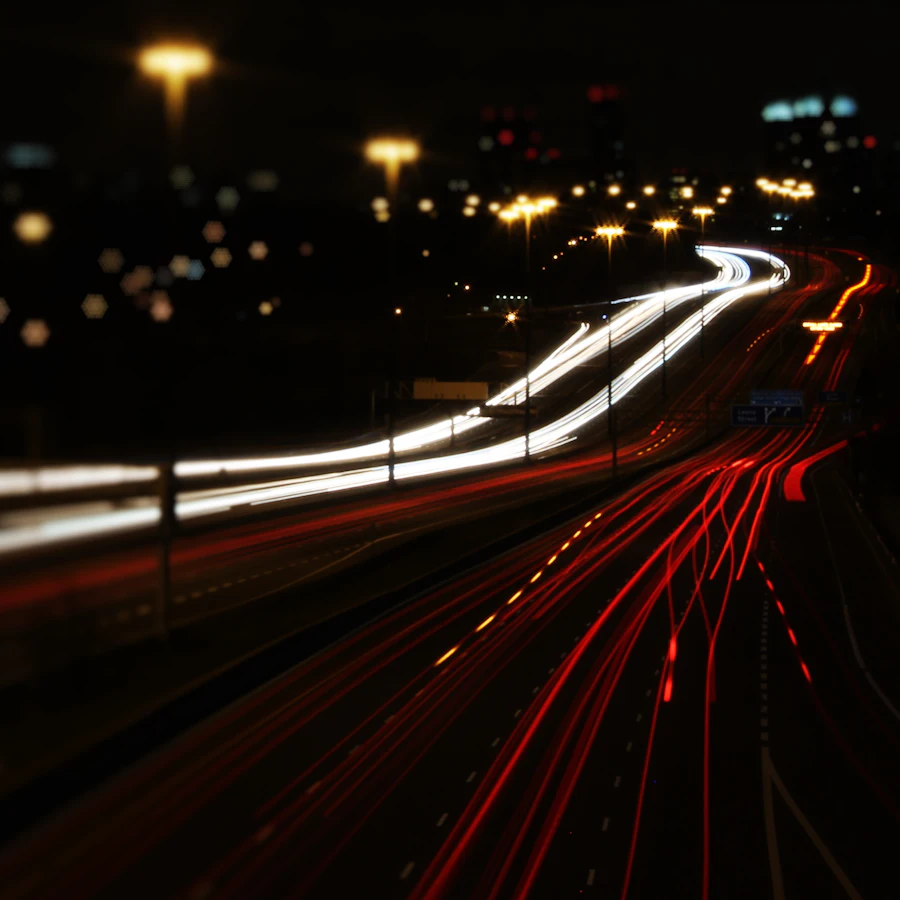 Long-exposure light trails curve along a dark highway at night, with white headlights and red taillights streaming into the distance. Soft city lights glow in the background.