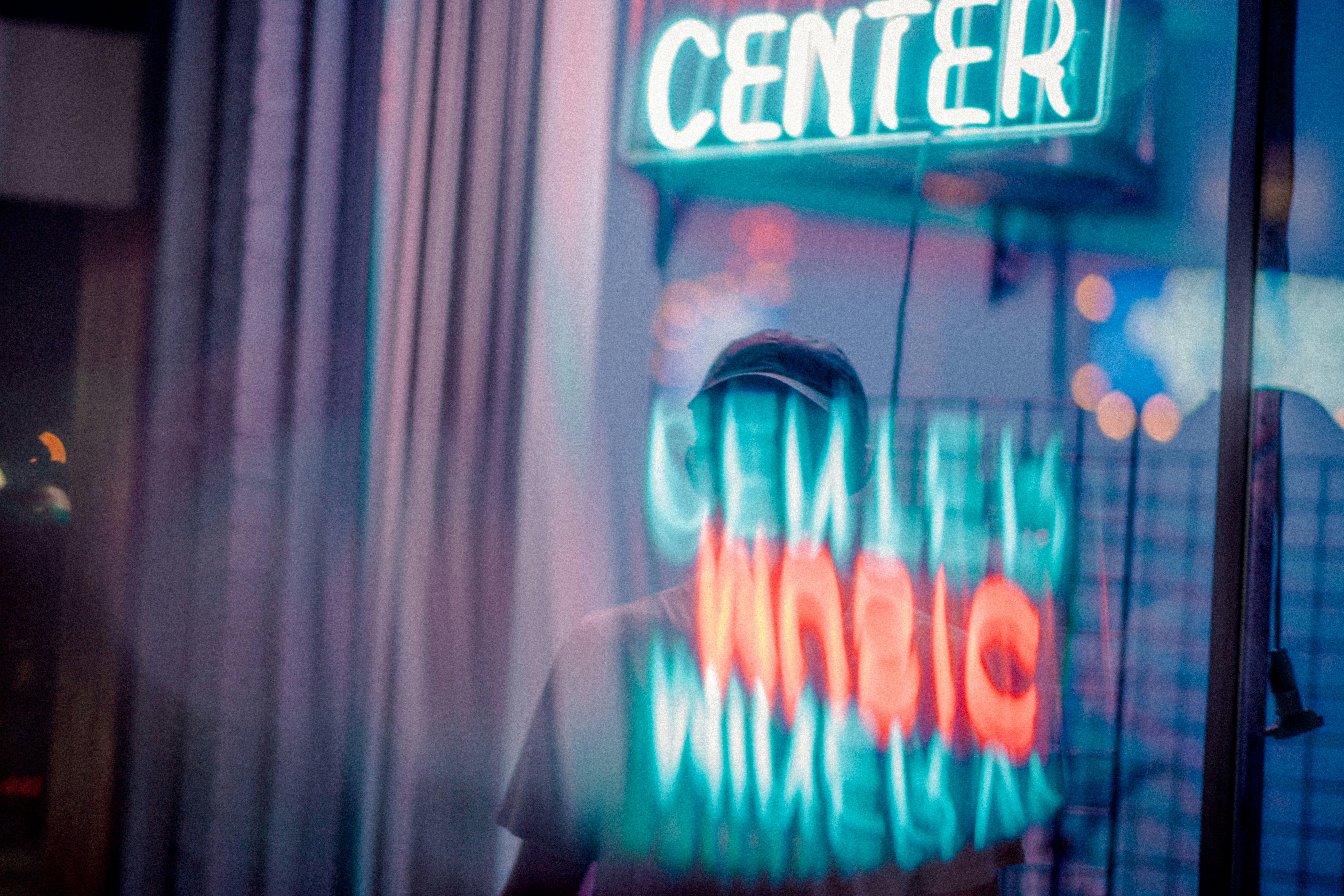 Blurred reflection of a person in a window with vibrant neon signage in blue and red hues.