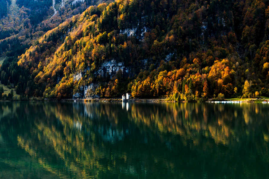Golden and green autumn forest covers a steep mountainside above a calm lake, with a small white building near the shore. The colorful trees and cliffs are mirrored in the still water.