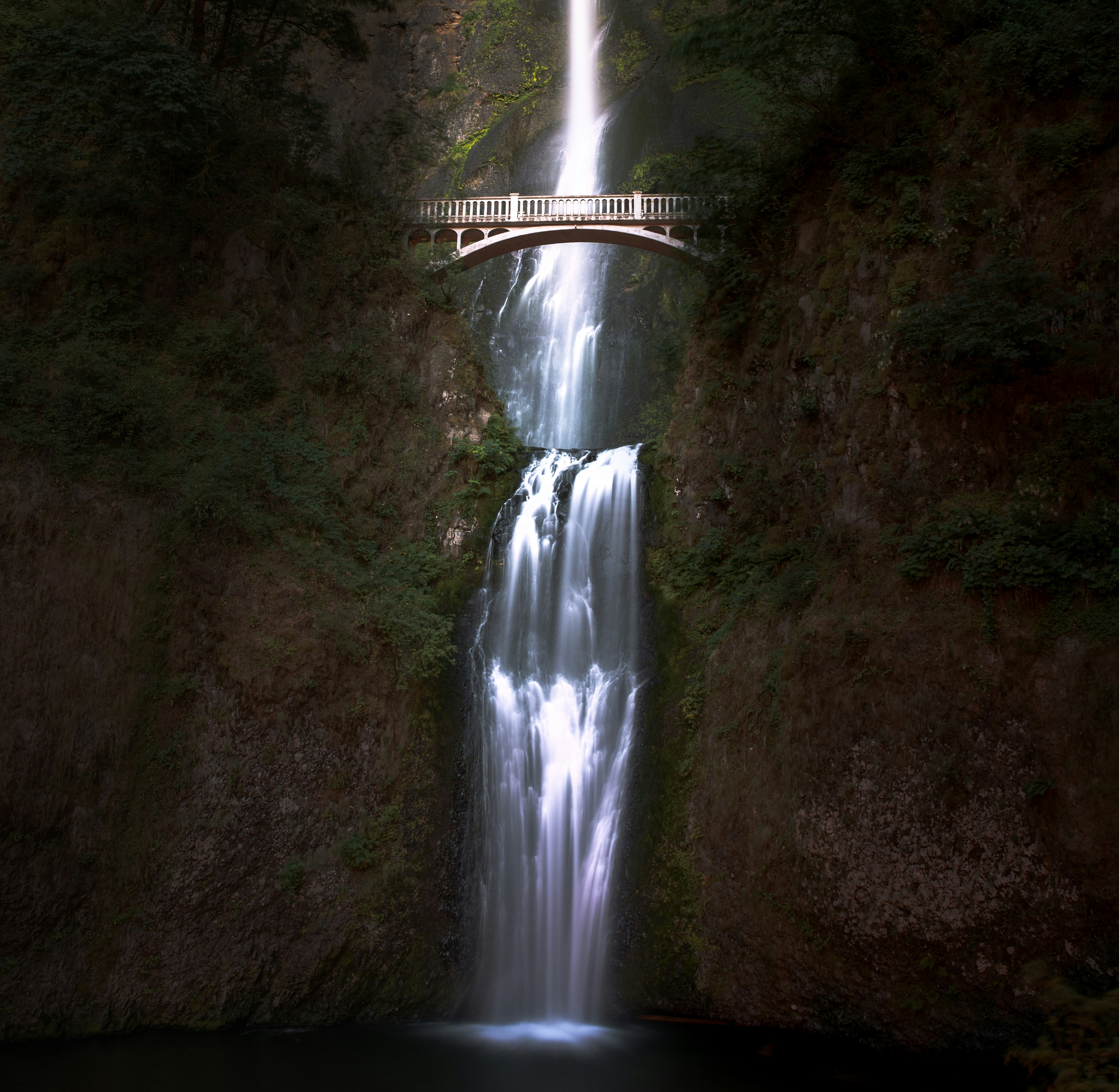 Majestic waterfall cascading beneath a historic bridge, surrounded by lush greenery and rocky cliffs.