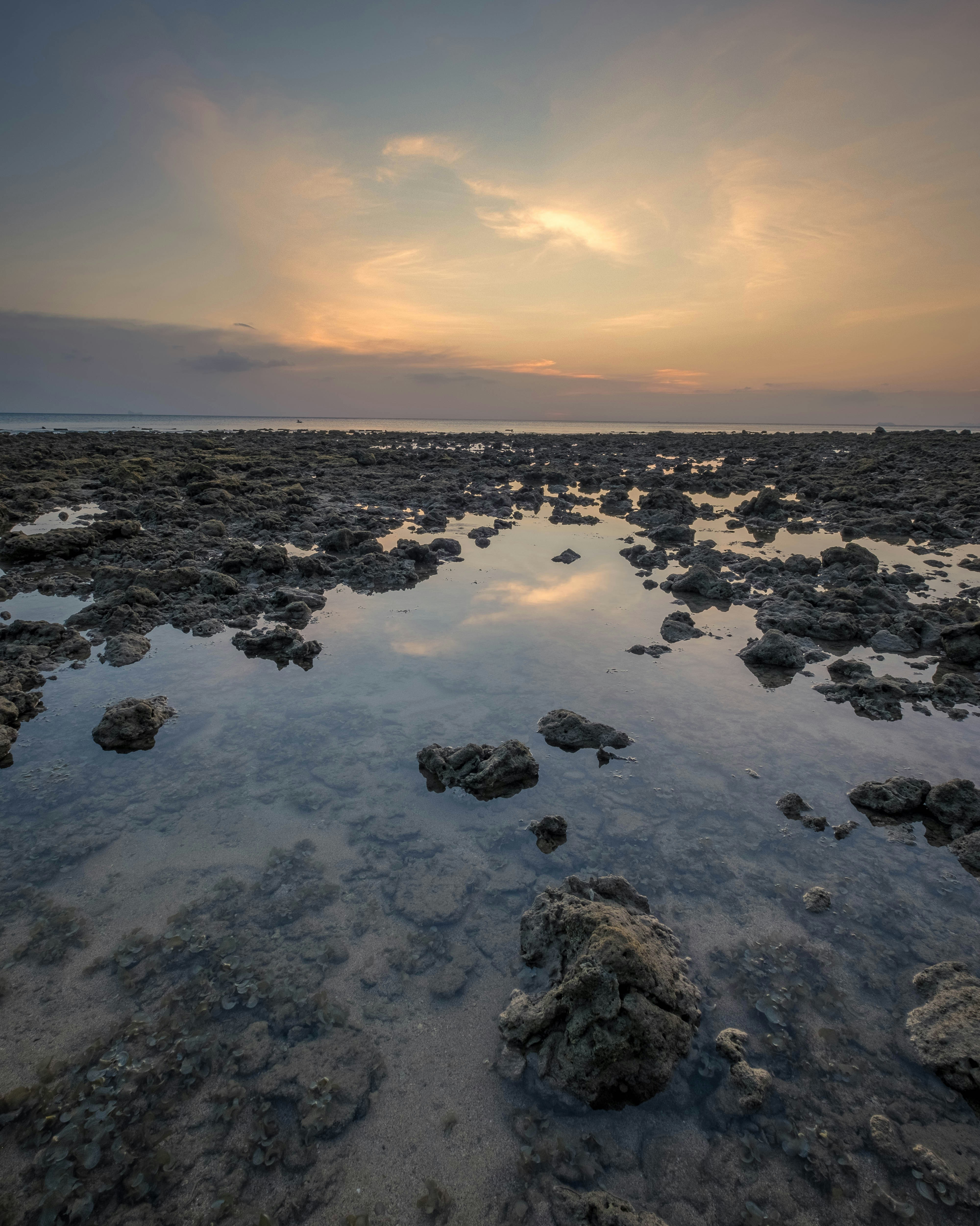 Rock formations and tidal pools reflect the soft hues of a sunset sky over a tranquil beach.