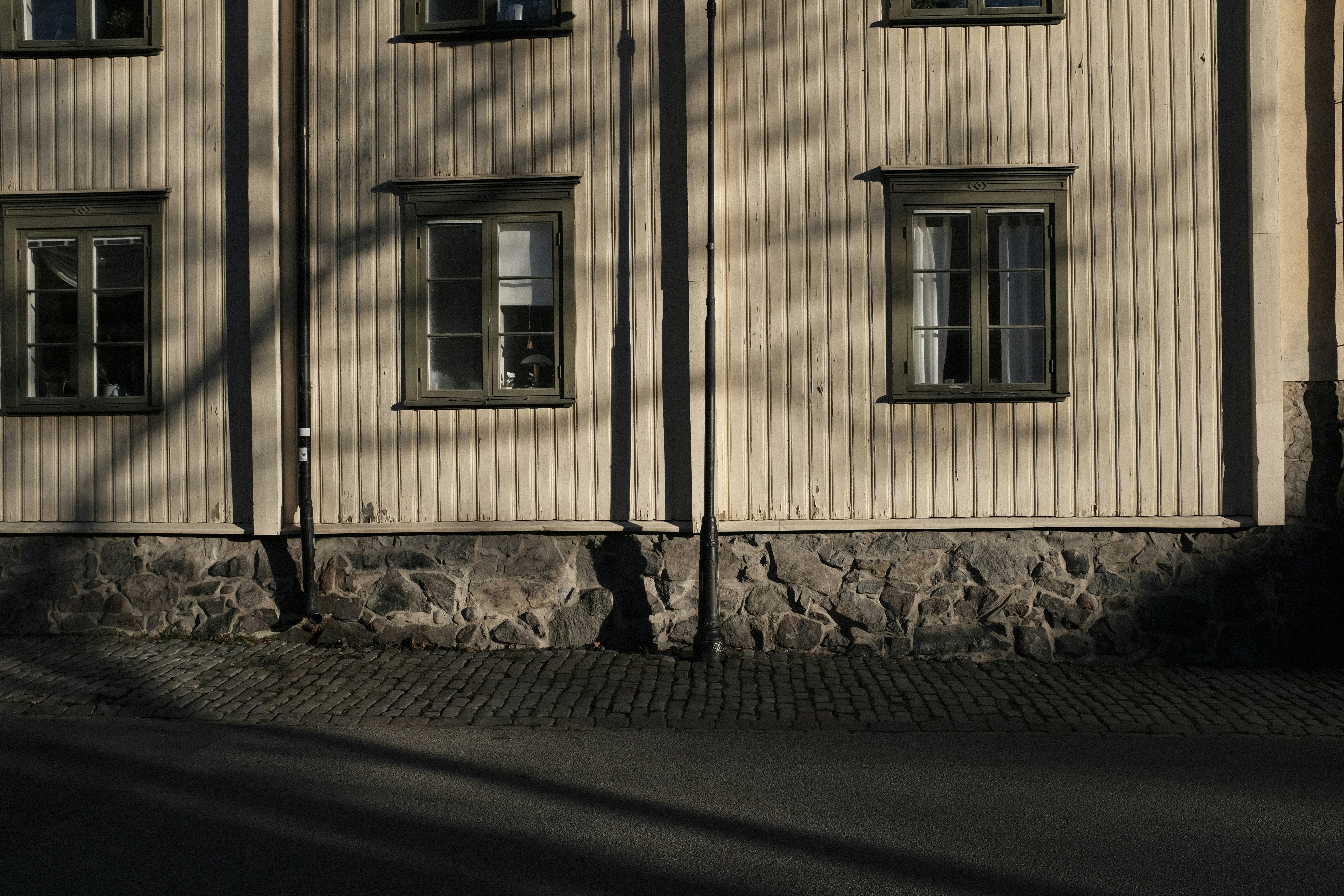 Warm-toned wooden building with symmetrical windows casting intricate shadows on the stone foundation. The play of light adds depth to the architectural details.