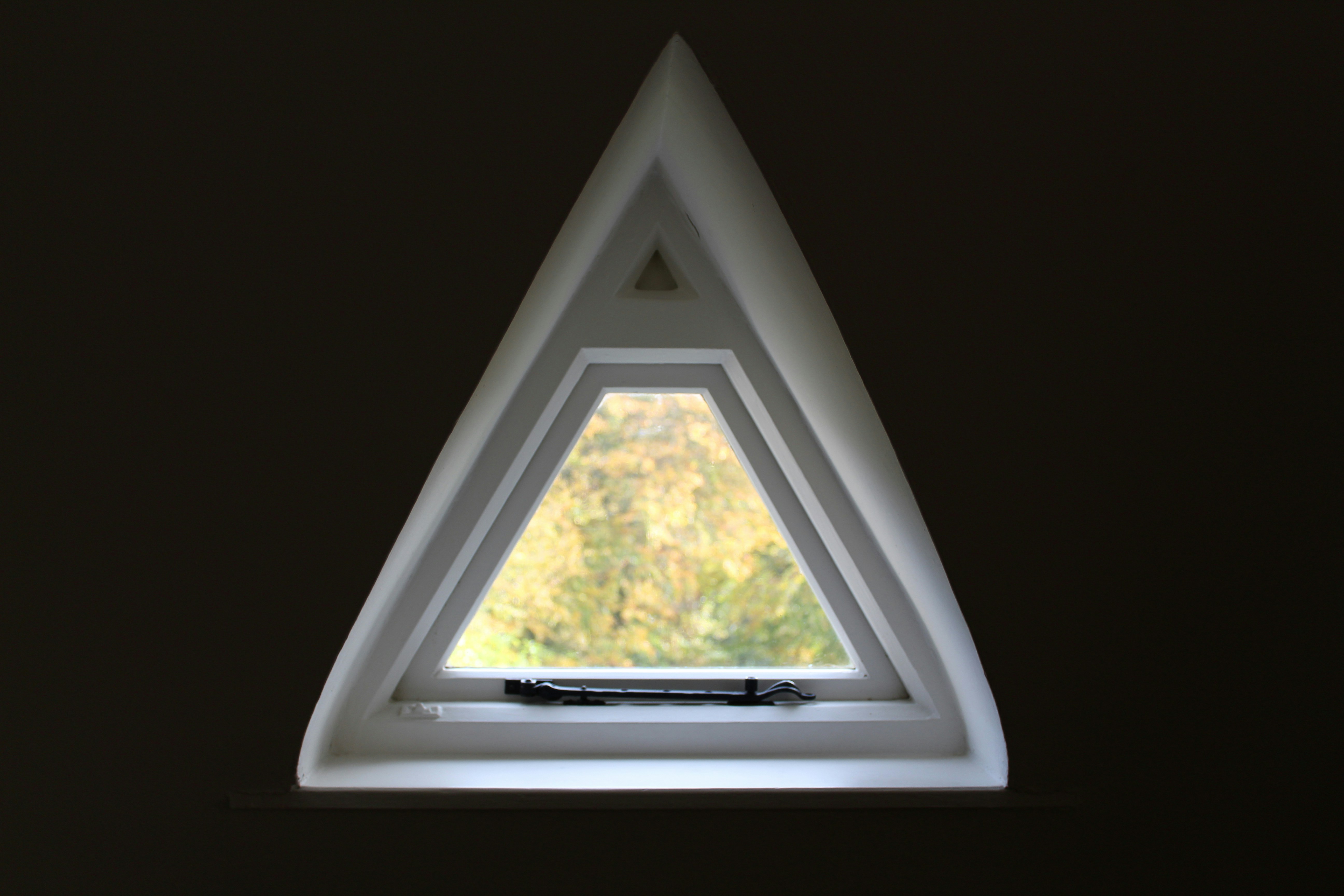 Triangular window with a white frame against a dark background, revealing a glimpse of autumn foliage.