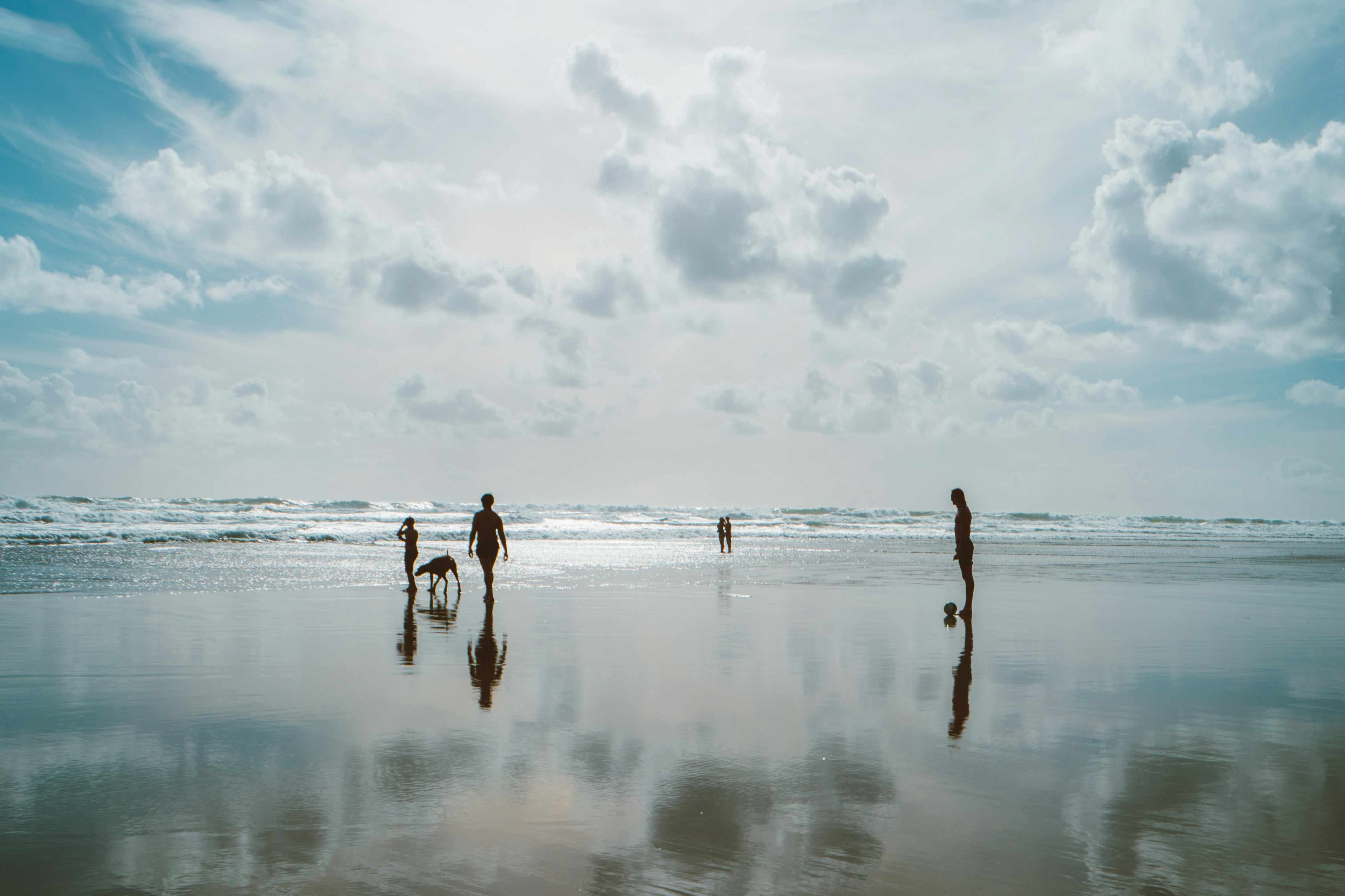 Silhouettes of people enjoying a beach day, with reflections shimmering on the wet sand under a cloudy sky.