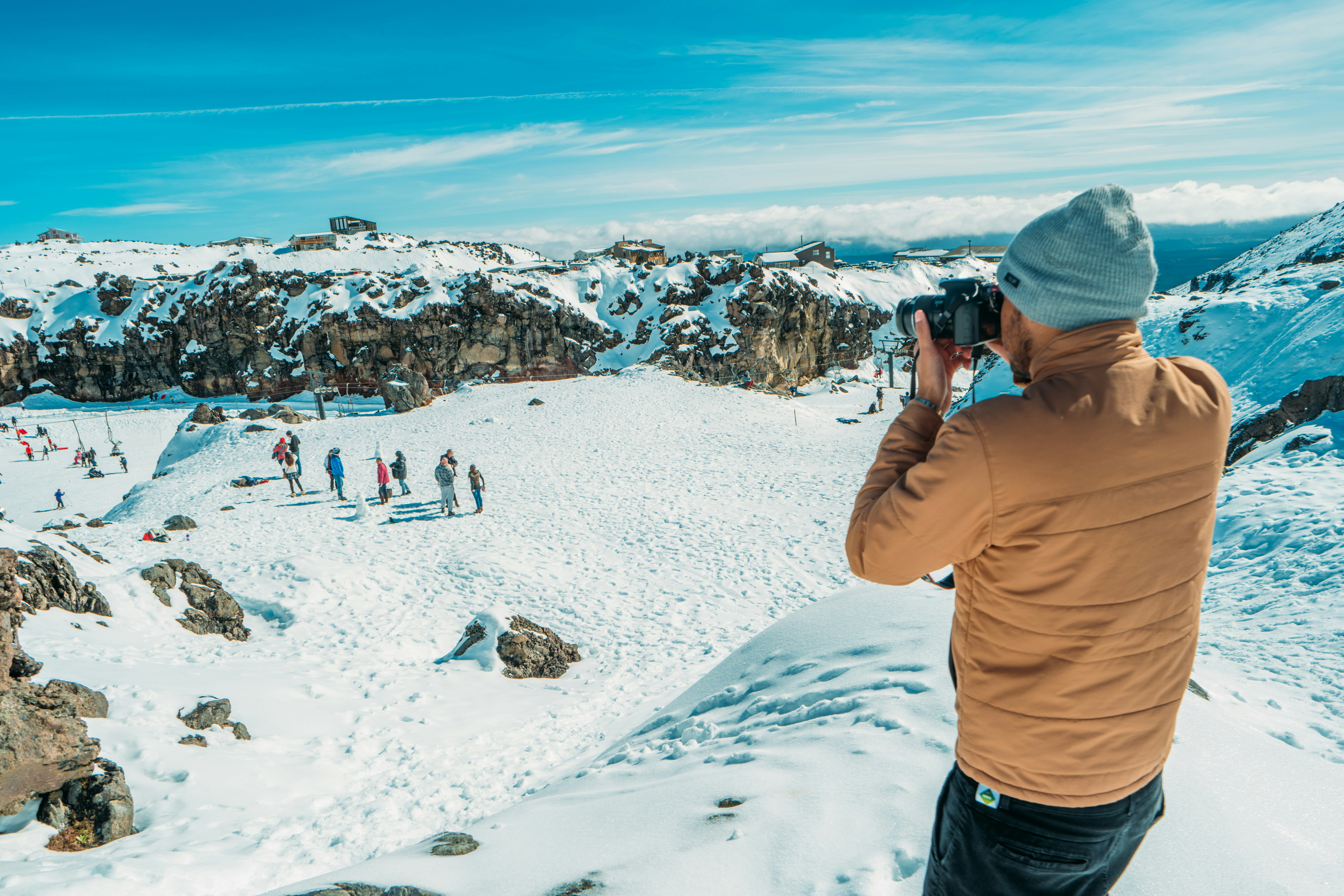 Person photographing snowy landscape with people in the distance under a clear blue sky.