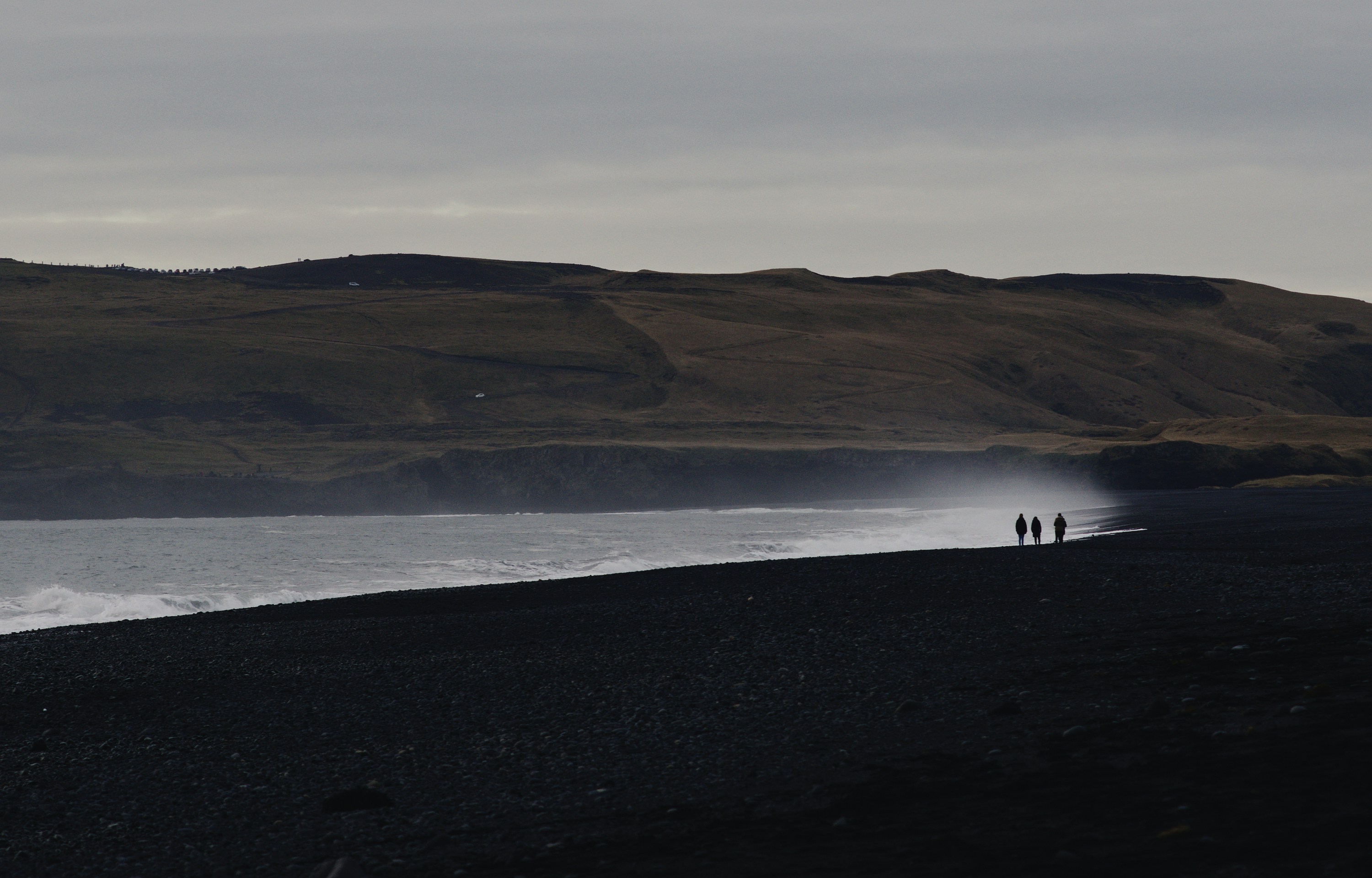 Moody coastal photograph of a dark pebble beach at dusk, with a distant group of walkers along the waterline under an overcast sky.