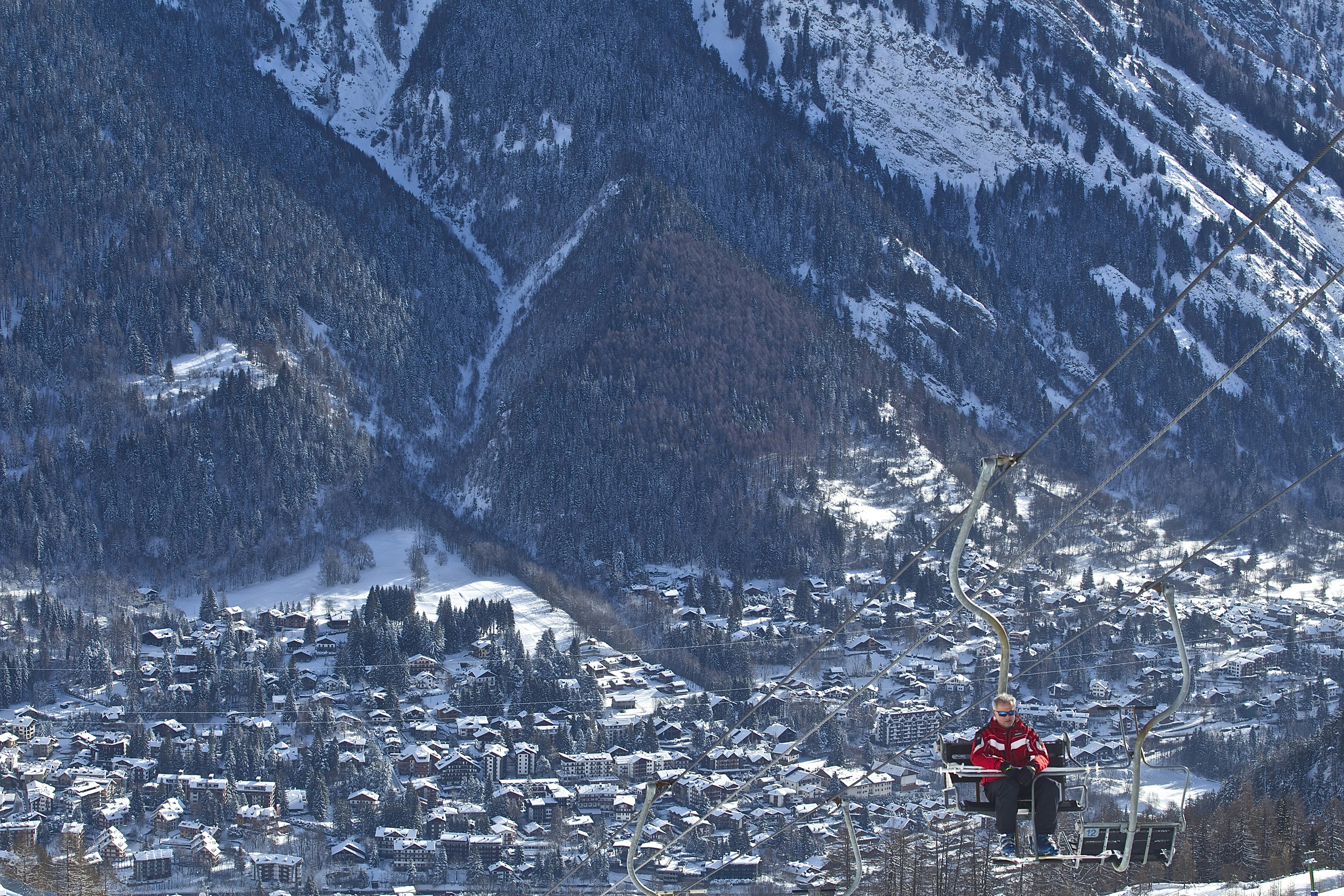Paraglider in red gear soaring over a snow-covered alpine village with majestic mountains in the background.