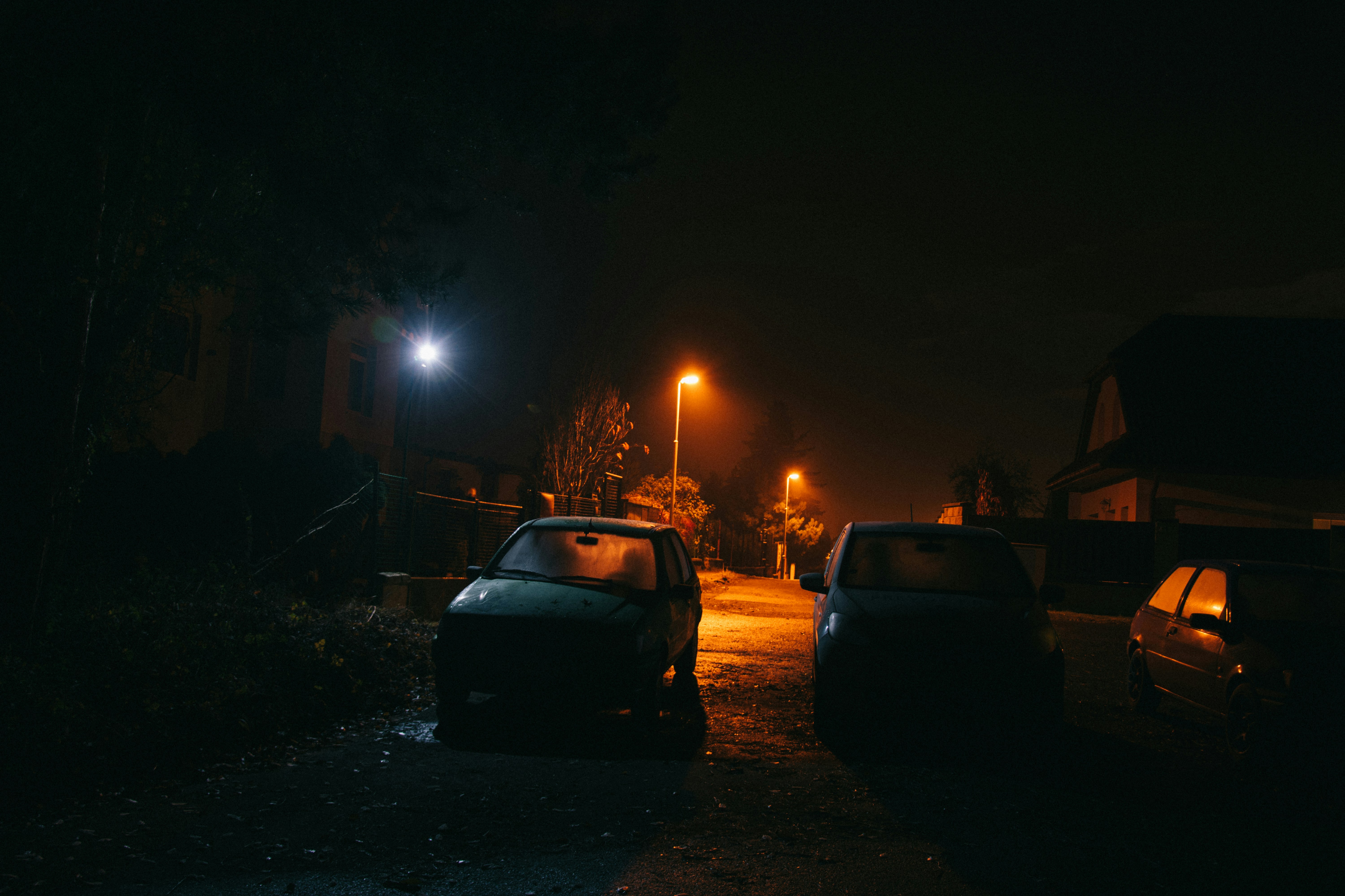 Two parked cars glisten under streetlights on a quiet, rainy night, surrounded by shadows and reflections.