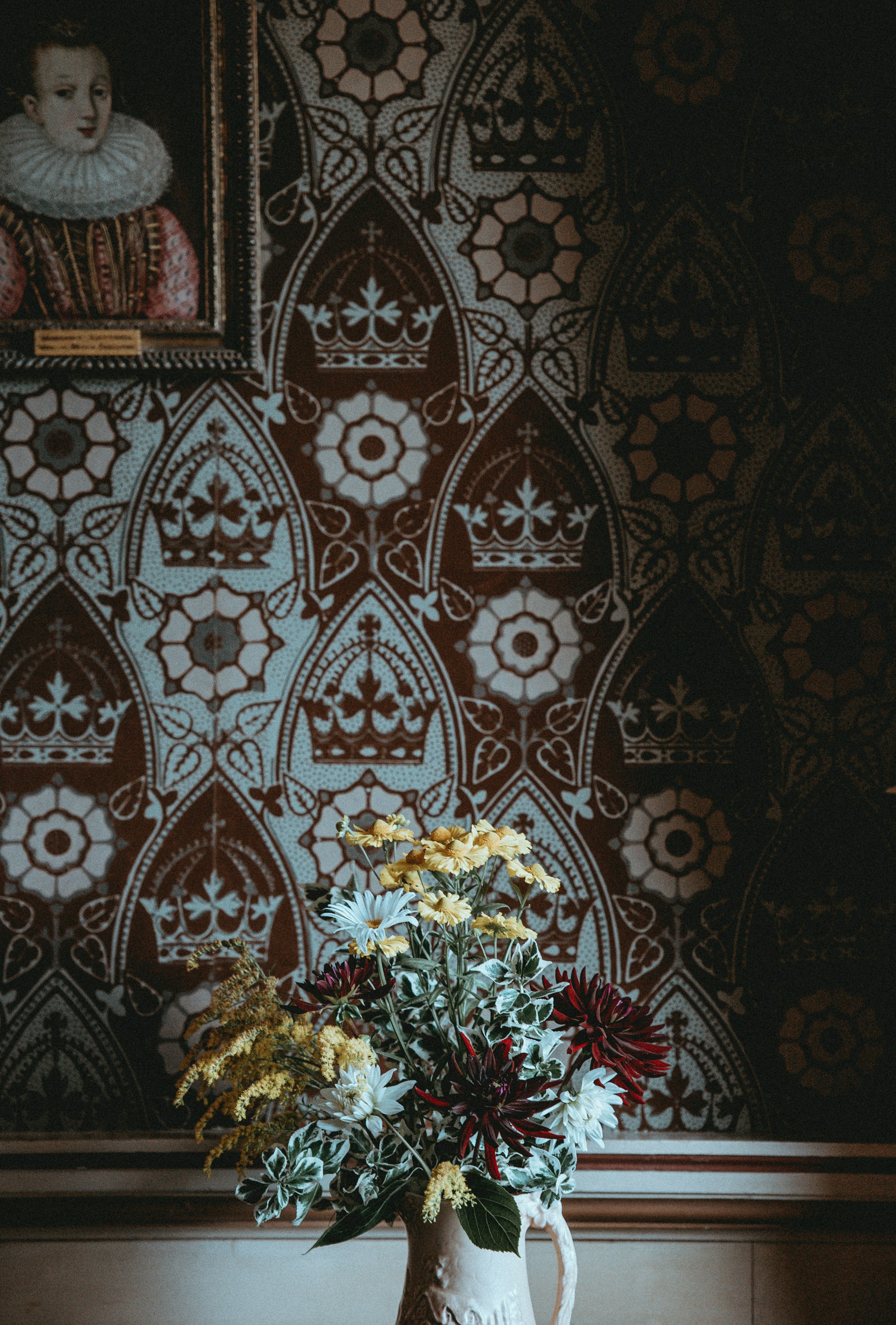 Vase filled with vibrant flowers set against a richly patterned wall, featuring a classic portrait above. The intricate design enhances the elegance of the scene.