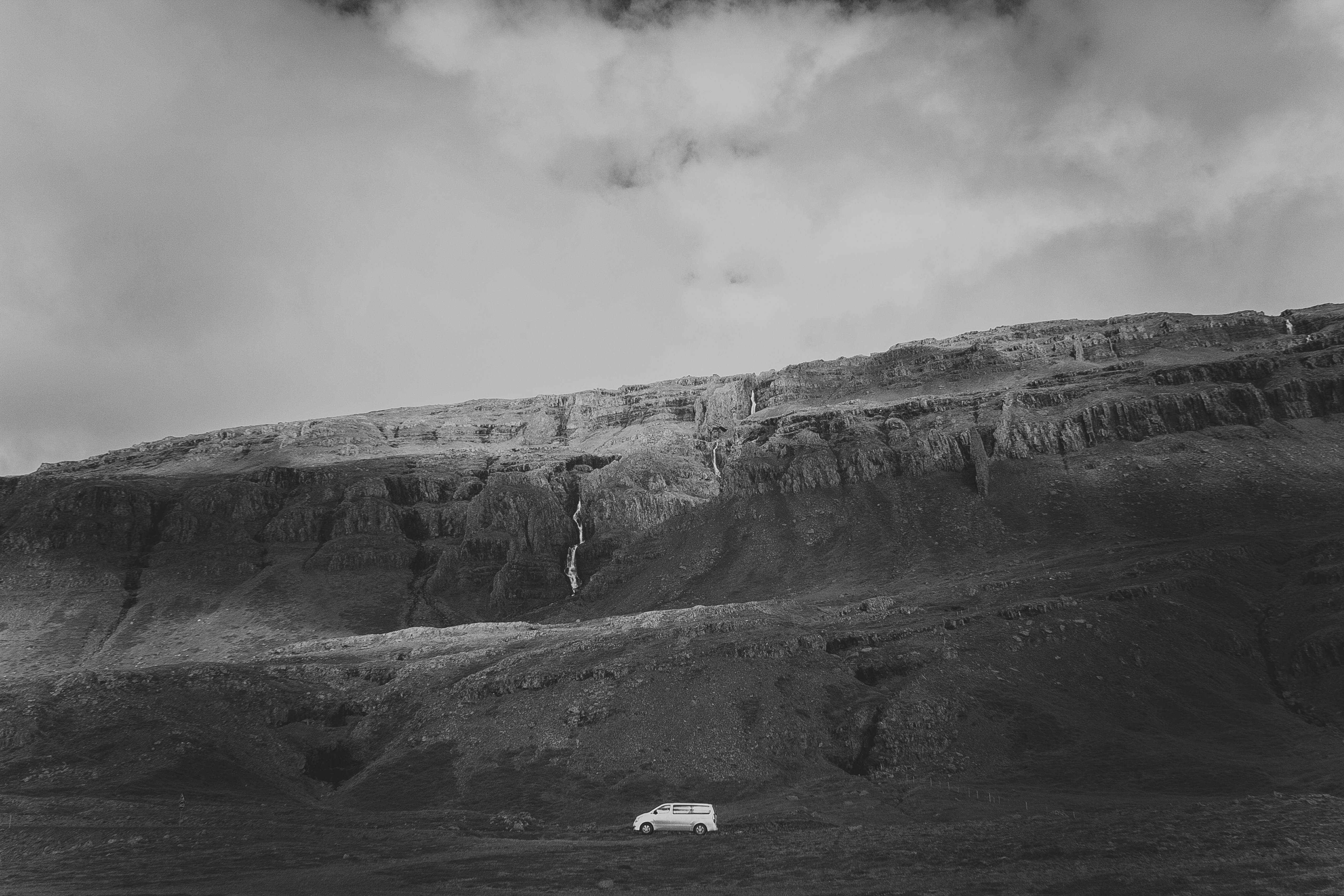 A lone van positioned against a dramatic, textured cliff face with cascading waterfalls, captured in black and white.