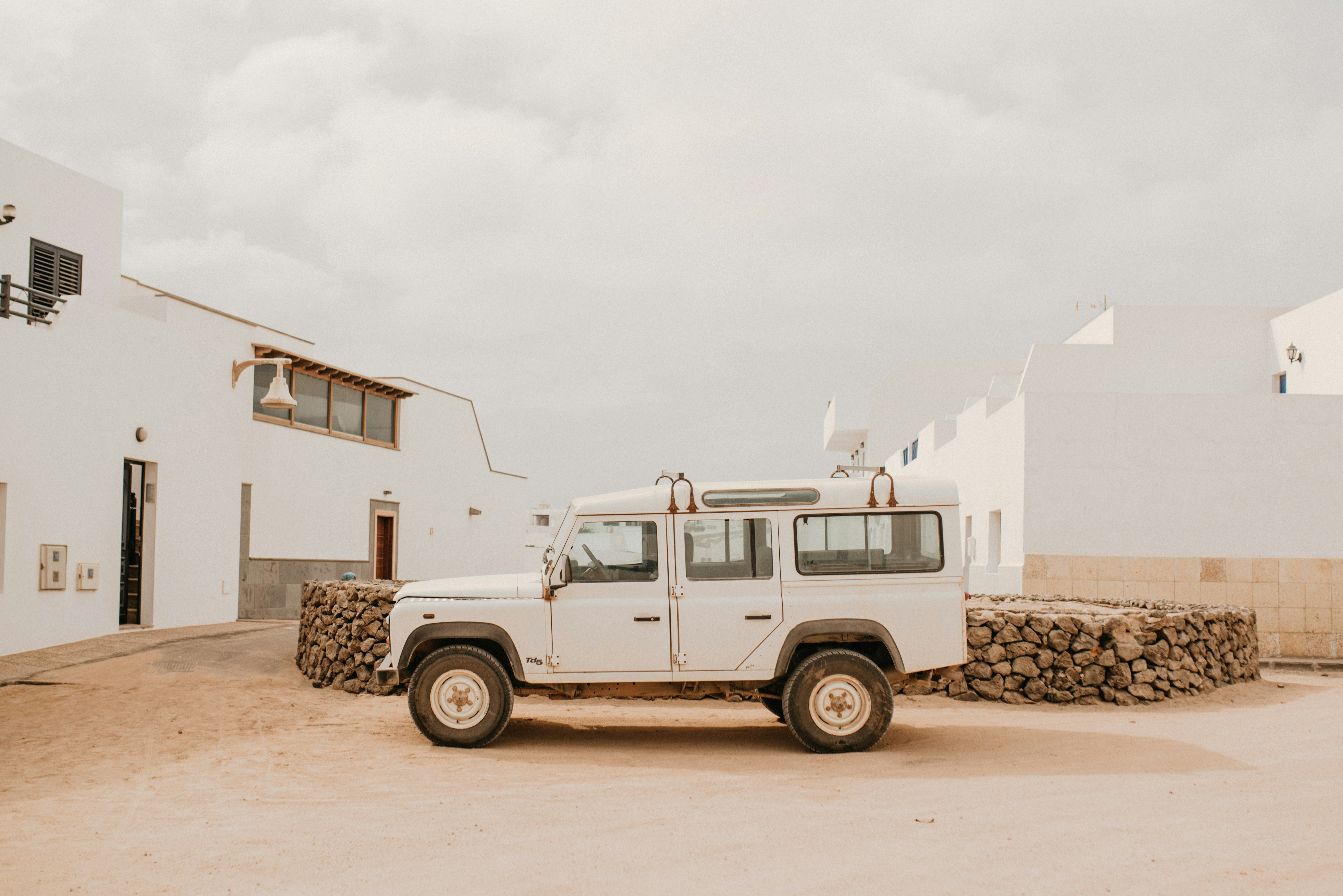 A vintage white Land Rover parked on a sandy road surrounded by minimalist white buildings and stone walls.