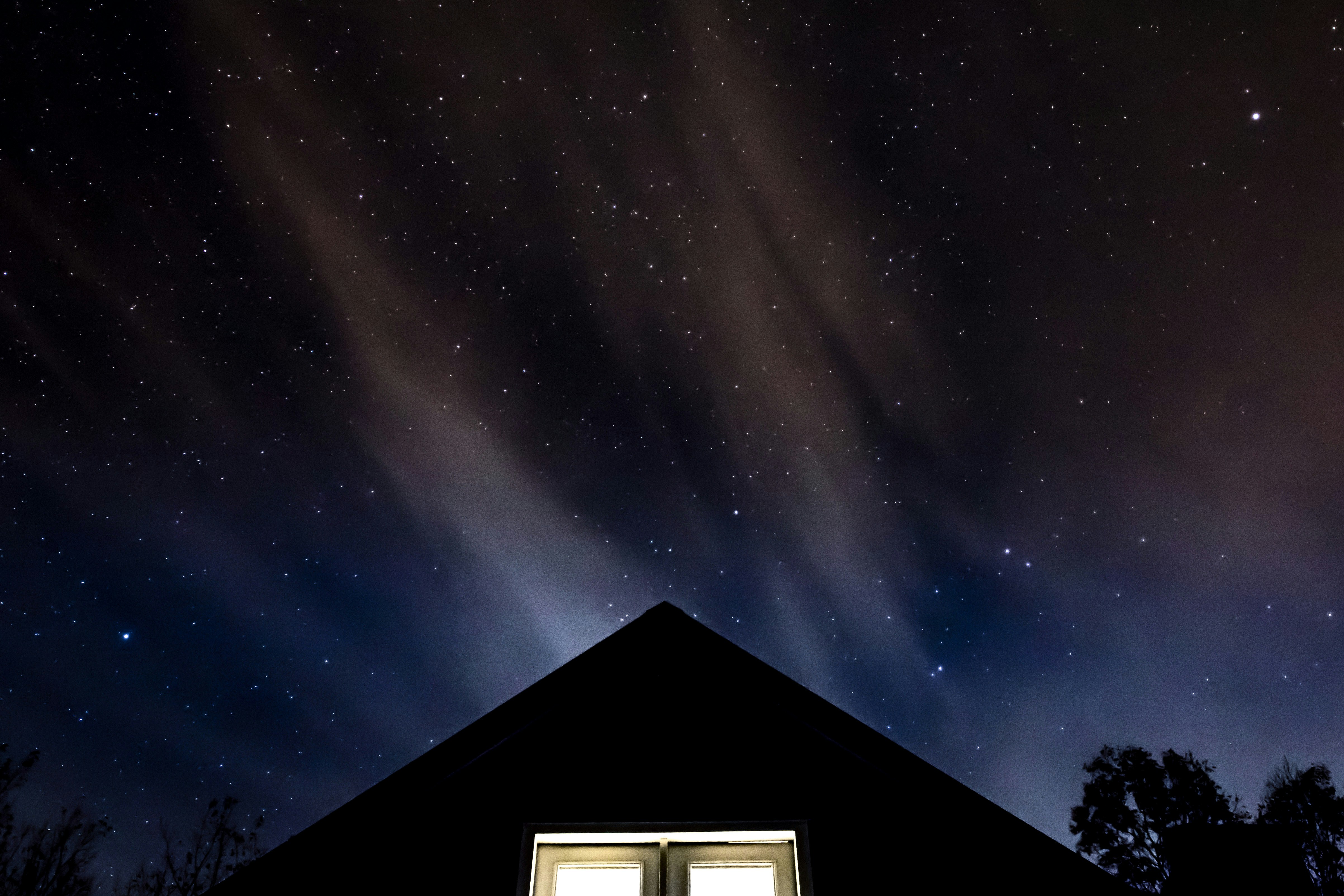 A silhouette of a house roof under a star-filled sky, with wispy clouds adding depth to the cosmic scene.