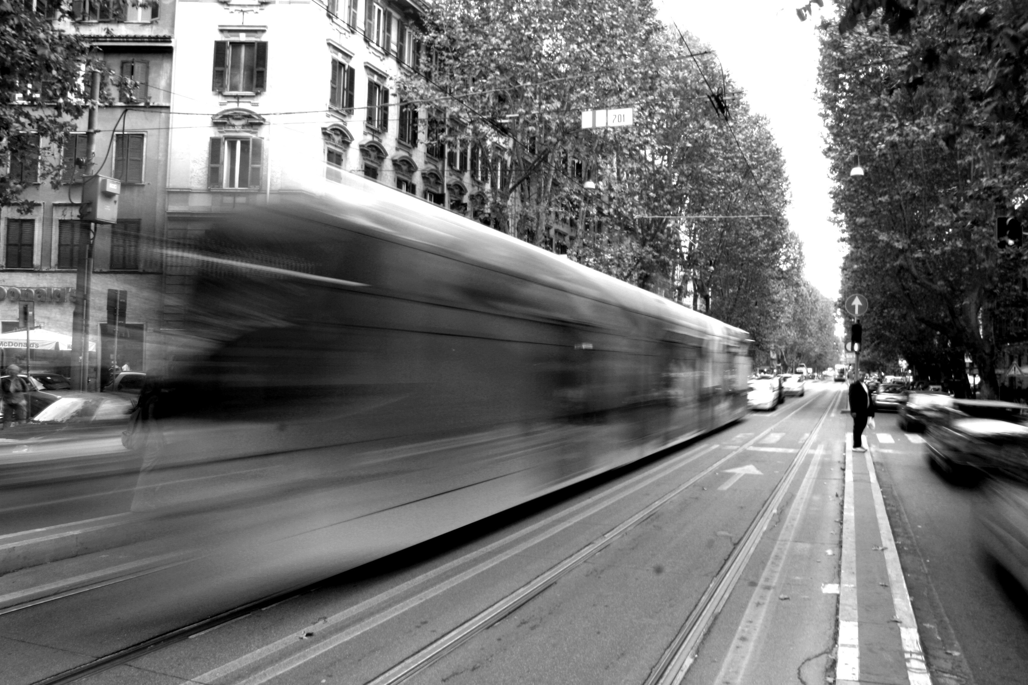 A blurred tram rushes past a lone figure on a city street, capturing the contrast between motion and tranquility in a bustling urban environment.