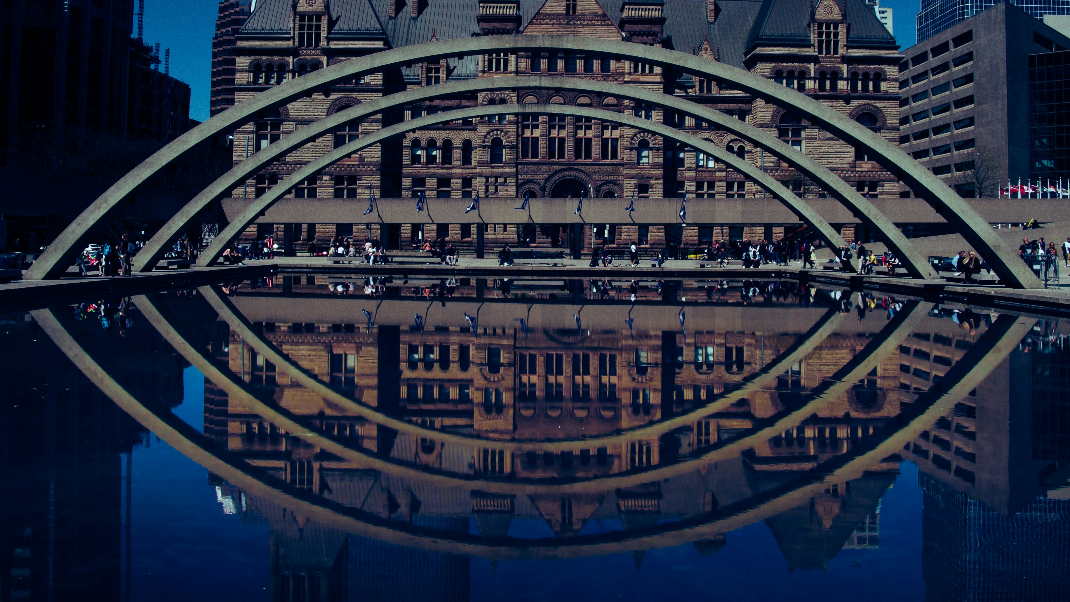 Historic building reflected in a serene water feature, framed by modern architectural arches. The scene captures a blend of old and new urban design.