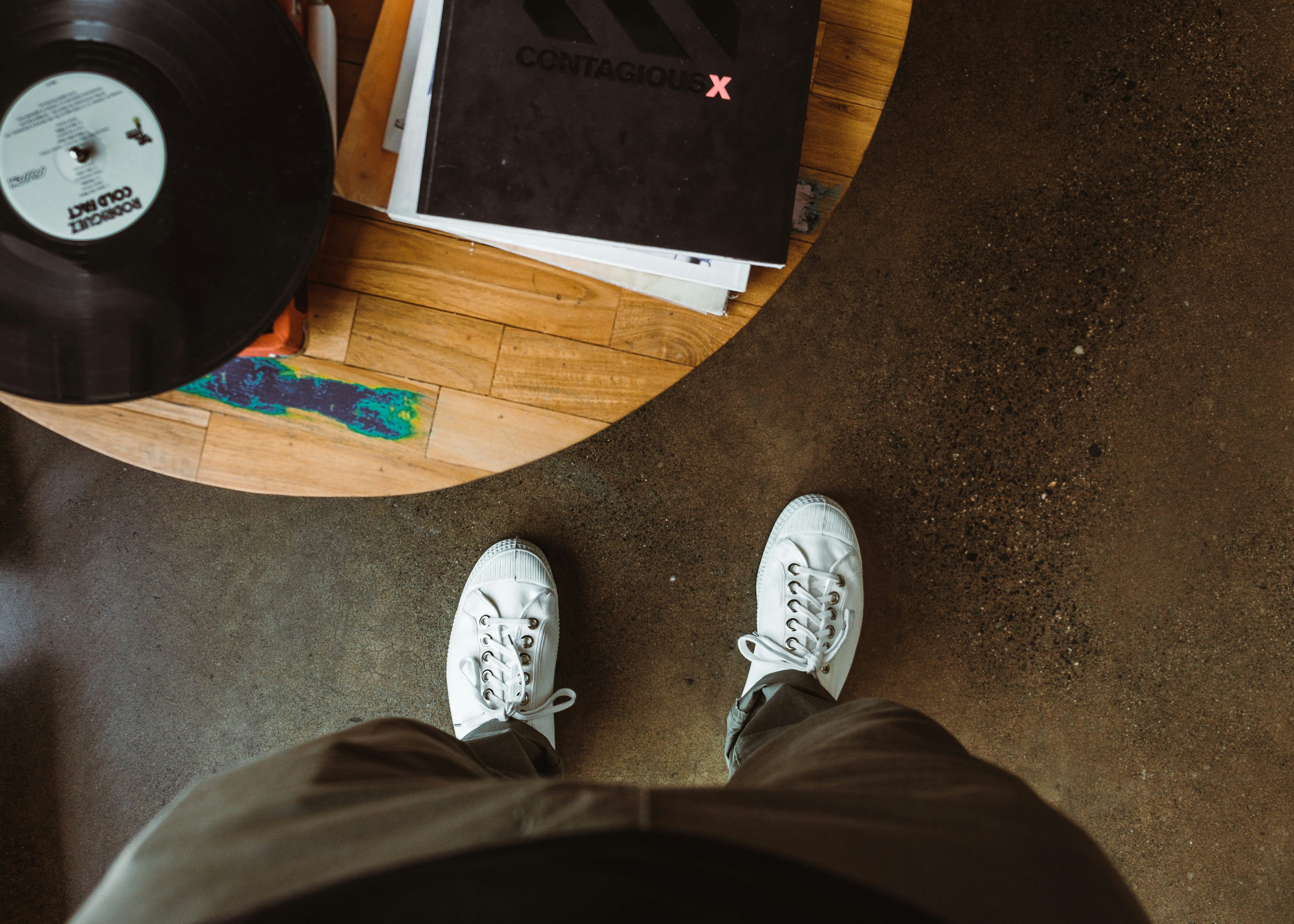 White sneakers on a concrete floor beside a round table with a vinyl record and a stack of books.