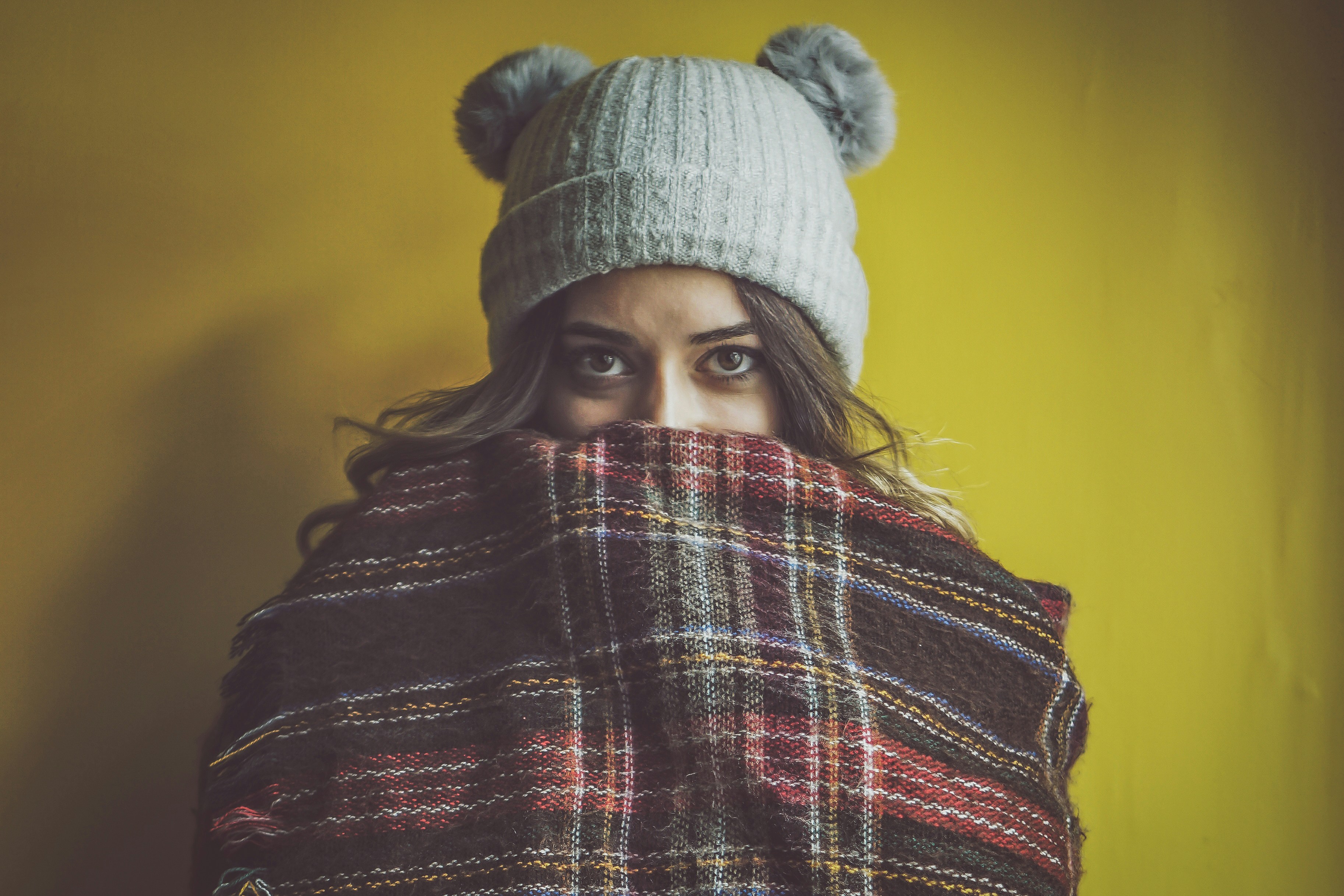Young woman wrapped in a colorful blanket, peering out from beneath a knitted hat against a vibrant yellow backdrop.