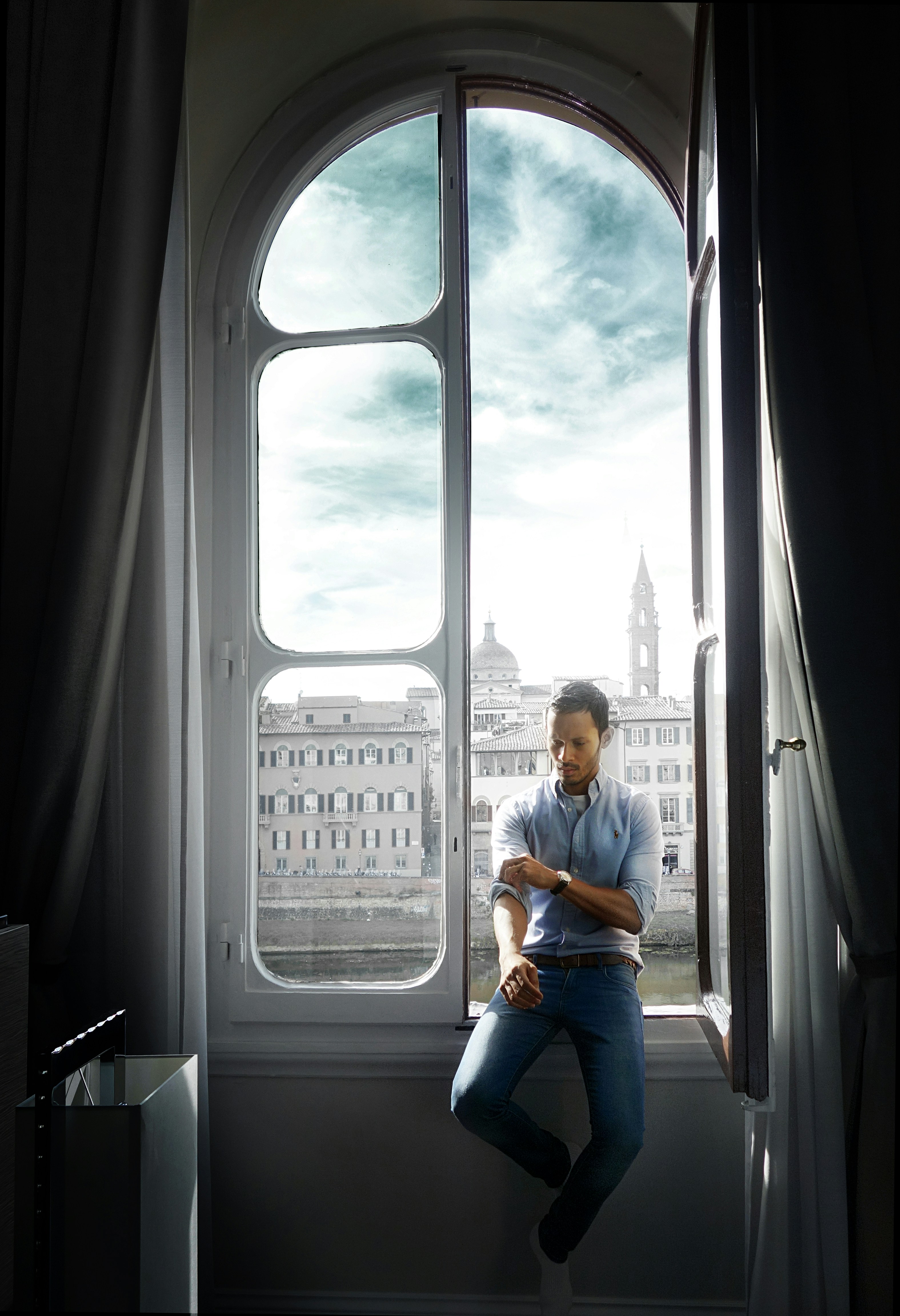 A man sits thoughtfully by a large window, overlooking a scenic cityscape with historical architecture. The soft light and clouds create a tranquil atmosphere.