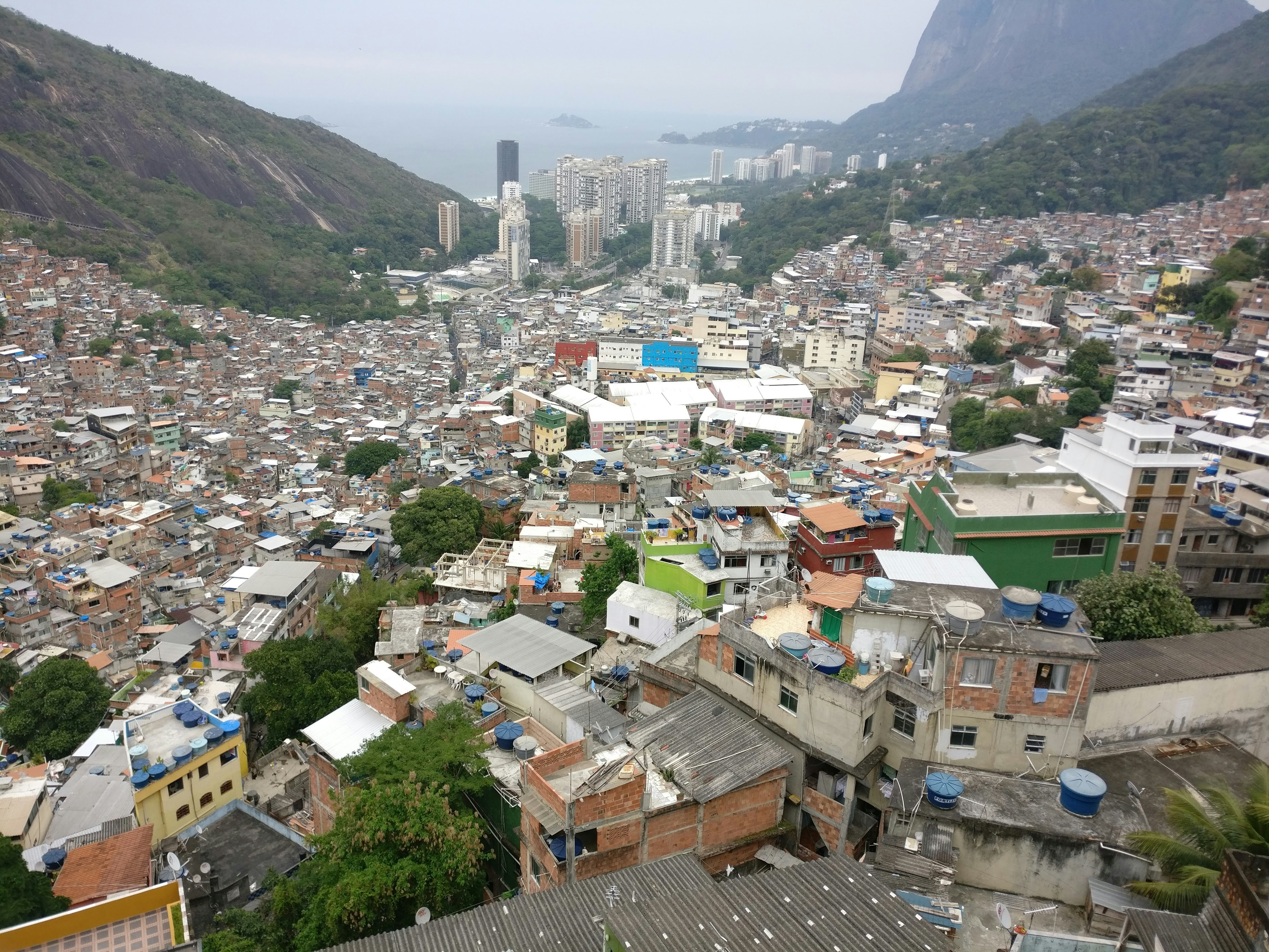 Panoramic view of densely packed buildings in Rocinha, Brazil, with distant hills and ocean visible under a cloudy sky.