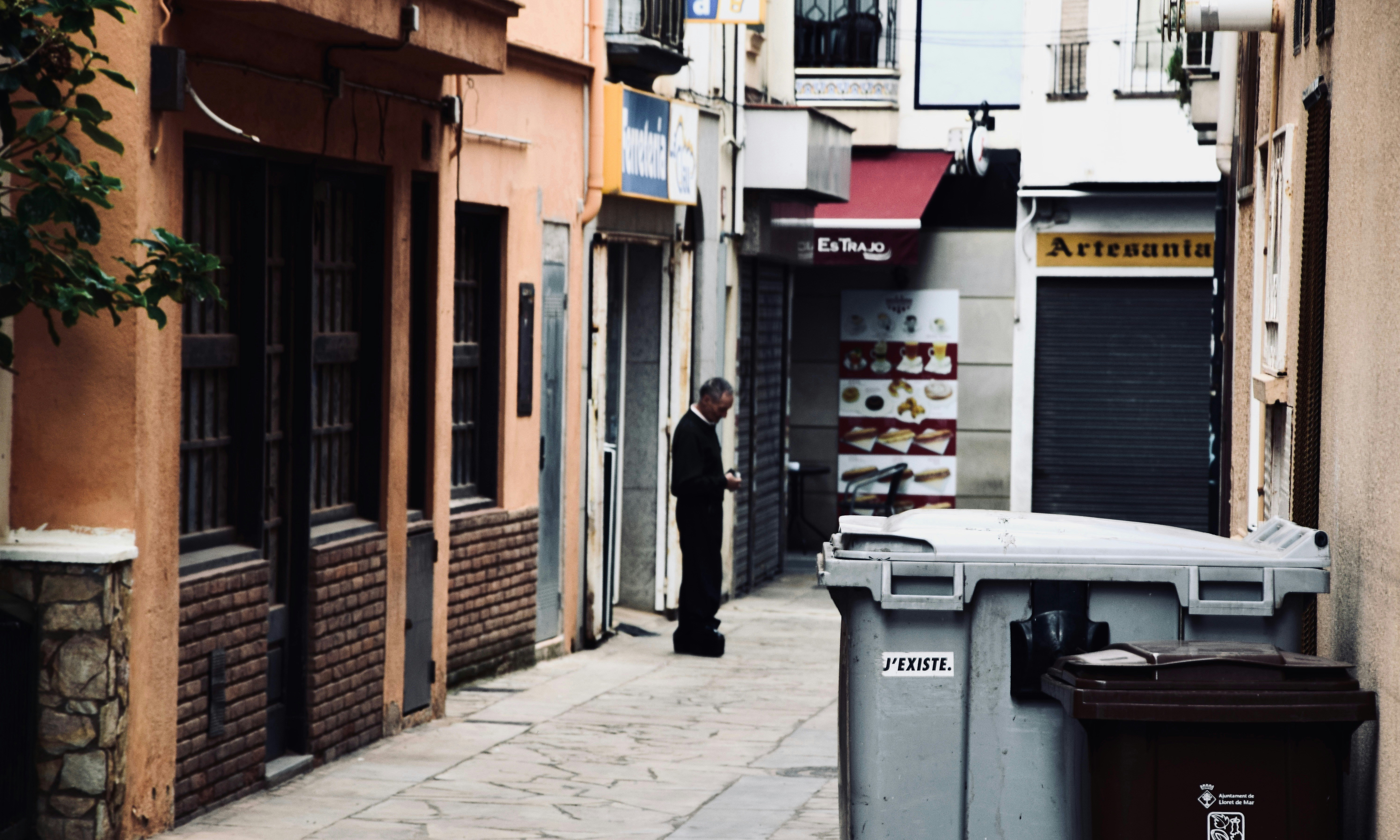 Person standing in a narrow alleyway with closed shutters and a garbage bin labeled 'J'existe.'.