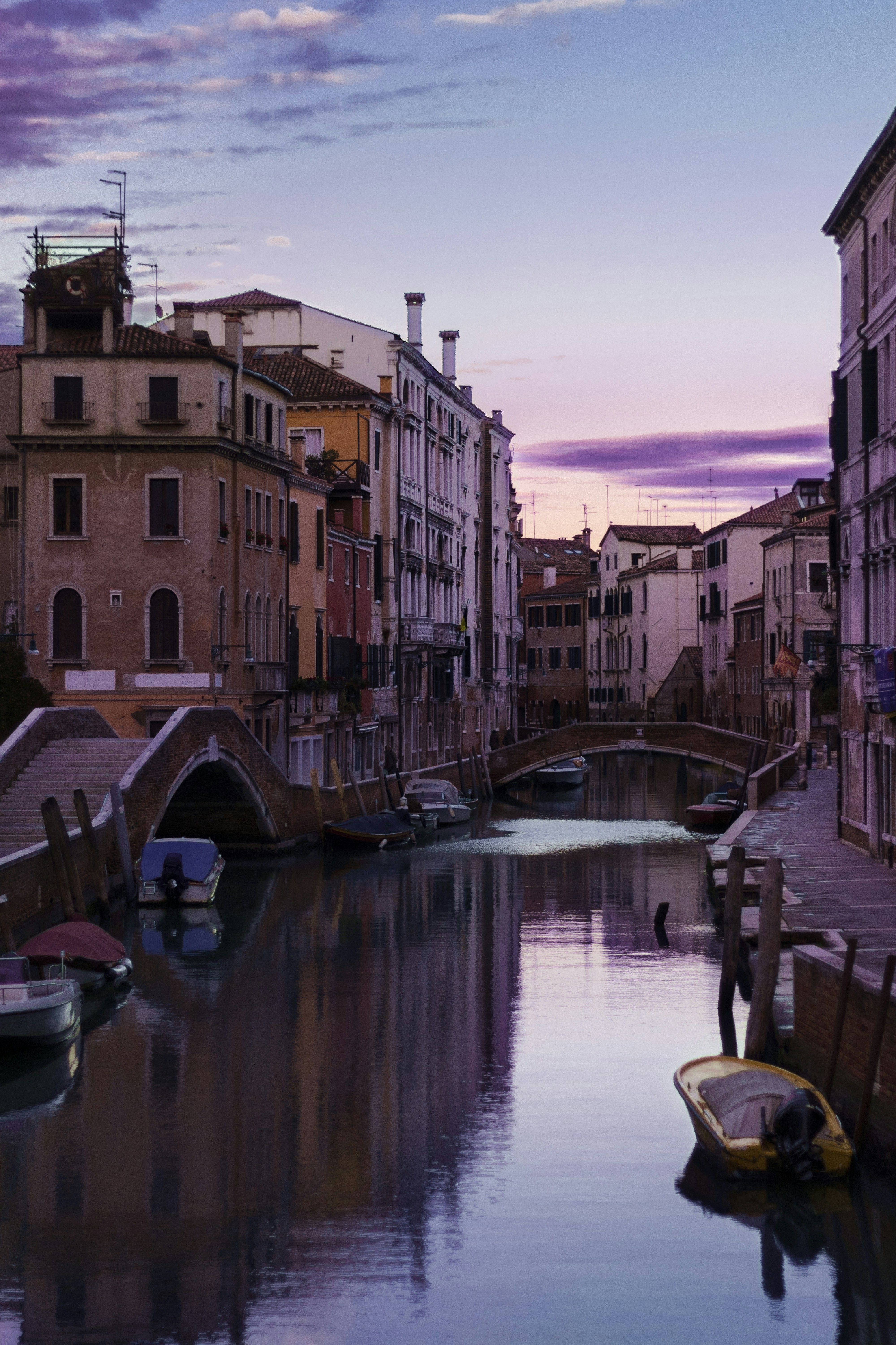 A serene canal in Venice reflecting twilight hues, framed by historic buildings and moored boats. The gentle arch of a bridge adds to the charm.