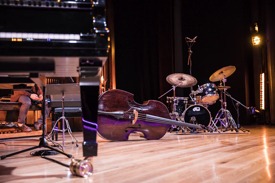 A double bass lies on a polished wooden stage in front of a drum kit, with stands and stage lights around it. A seated musician is partly visible at the left edge in the dimly lit venue.